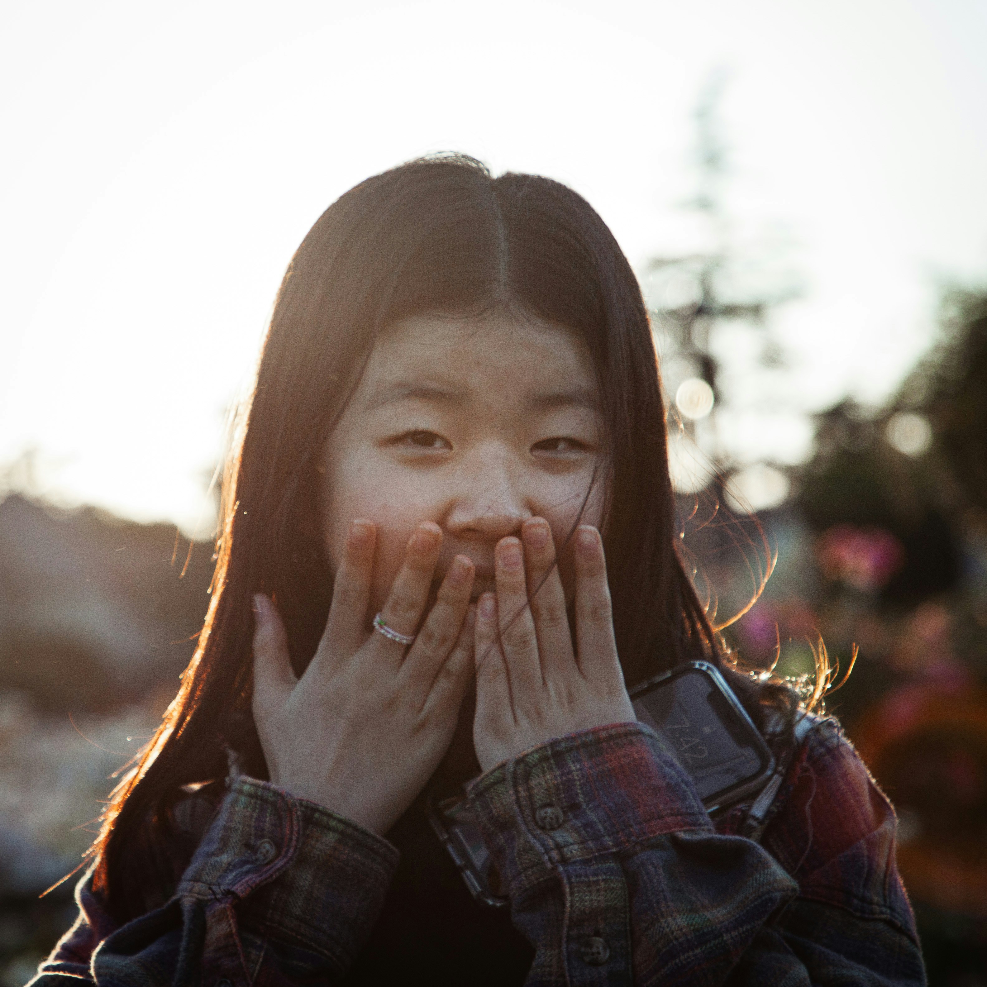 A girl playfully covering her mouth with her hands, surrounded by soft, warm light and blurred floral elements in the background.