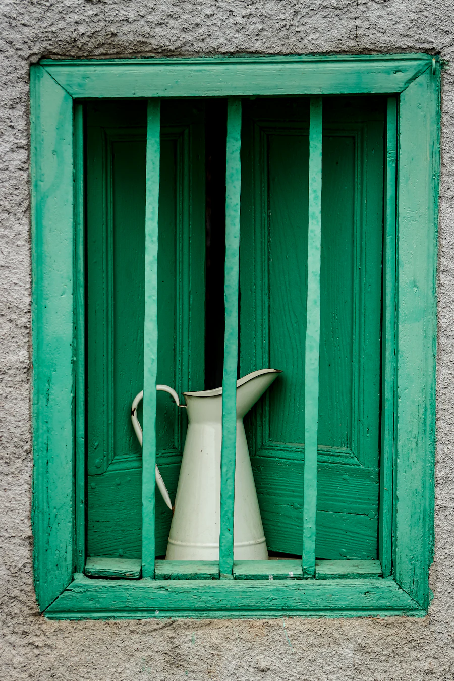 White enamel pitcher sitting behind vertical green bars in a weathered green window frame set into a rough gray wall.