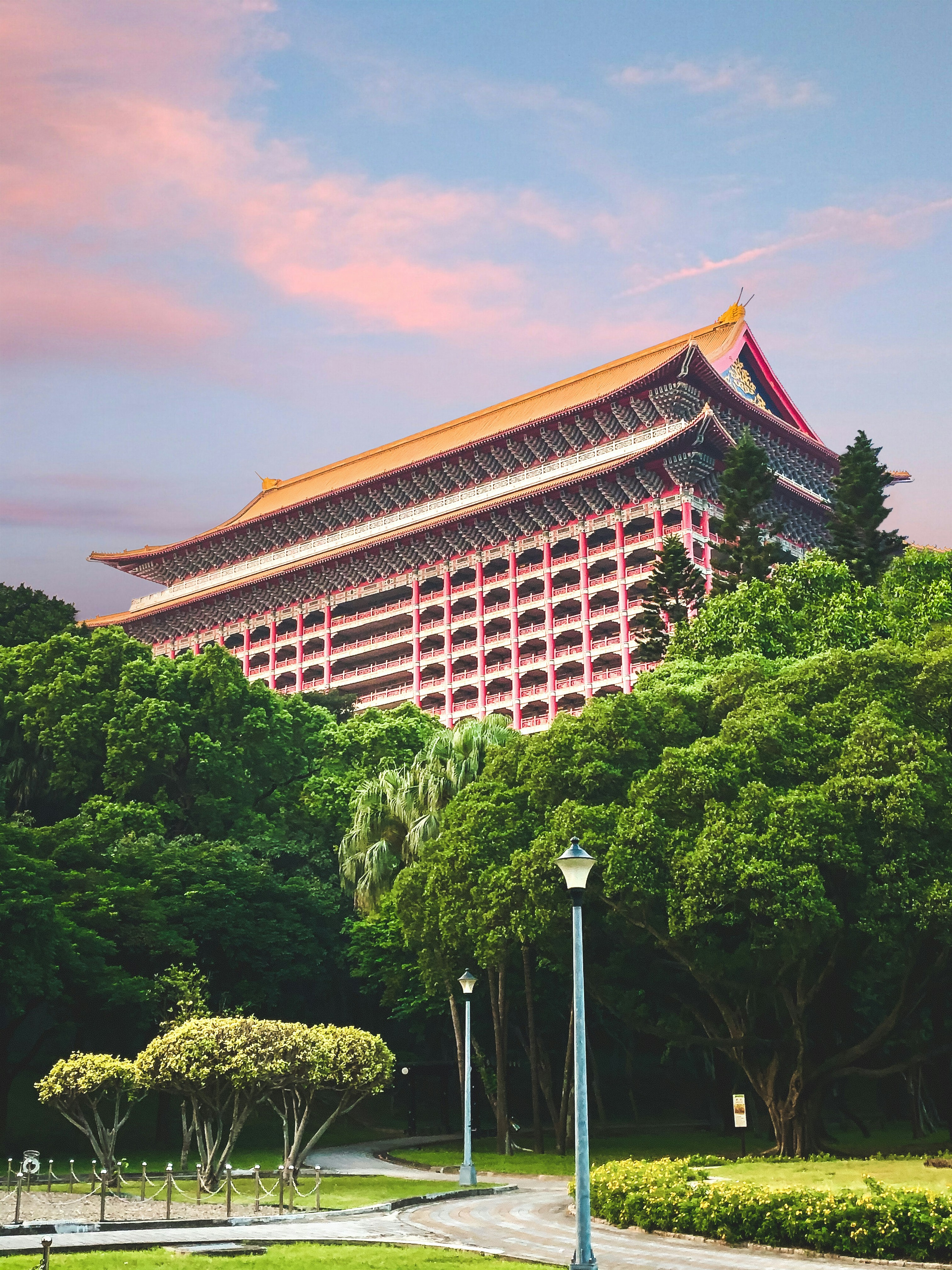 A grand traditional building rises above lush greenery, framed by a pastel sky at dusk.