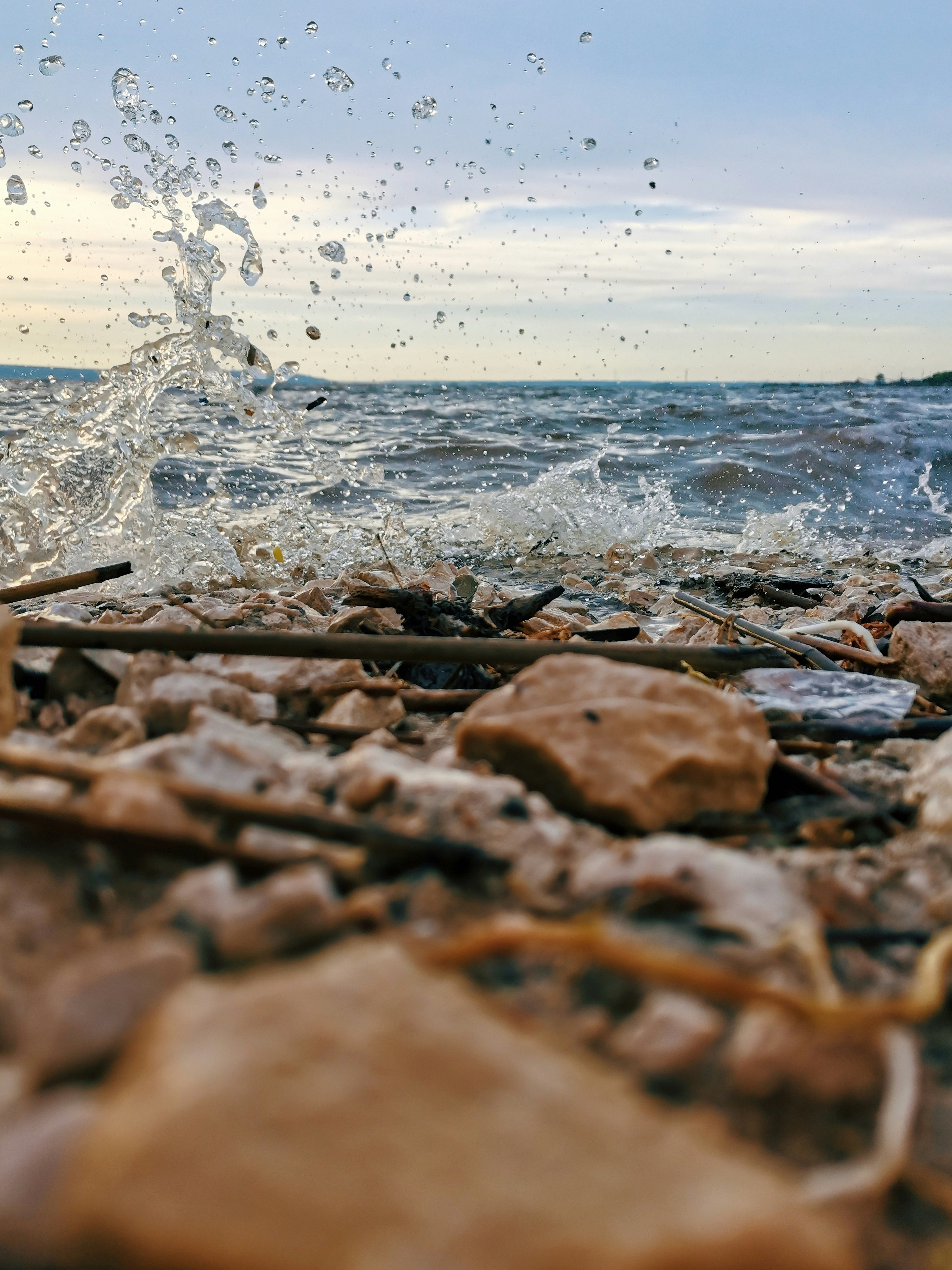 Water droplets erupting from the shoreline, surrounded by a mosaic of pebbles and twigs. The scene captures the dynamic interaction between land and water.
