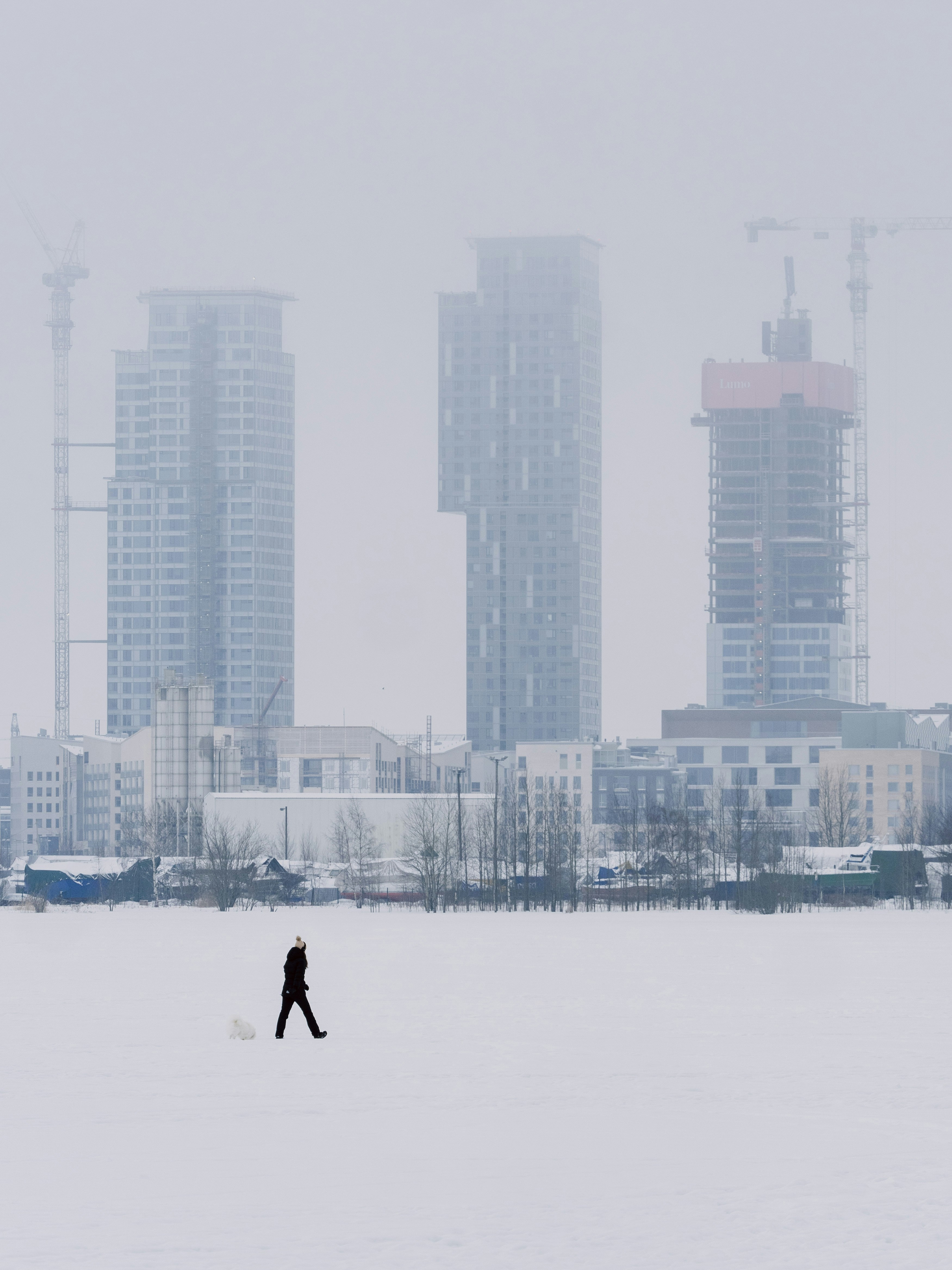 A lone figure walks on a snow-covered landscape, framed by towering construction cranes and modern buildings in the background.