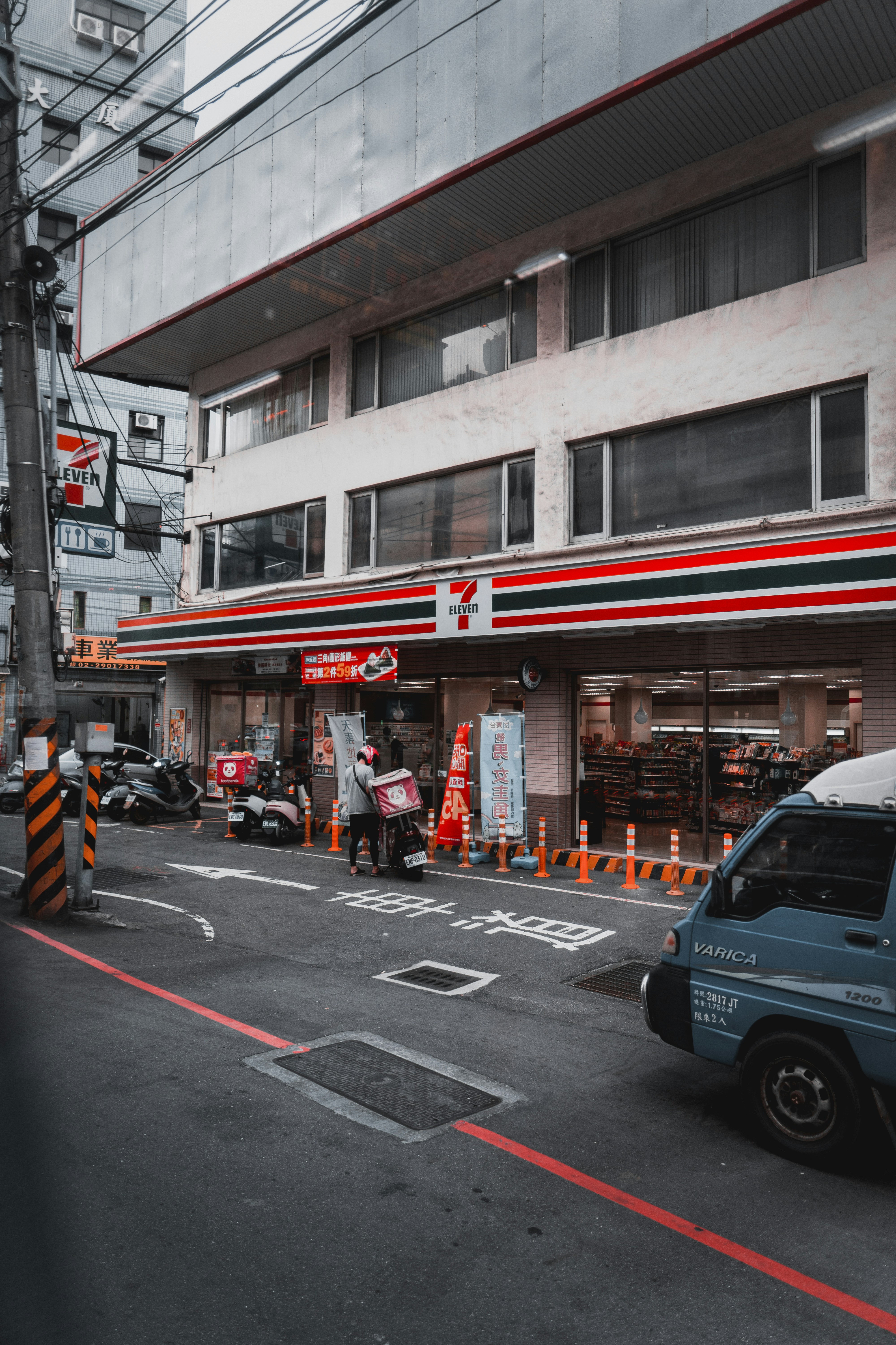 A bustling street scene featuring a 7-Eleven store with delivery personnel and parked scooters, showcasing the vibrancy of urban life.