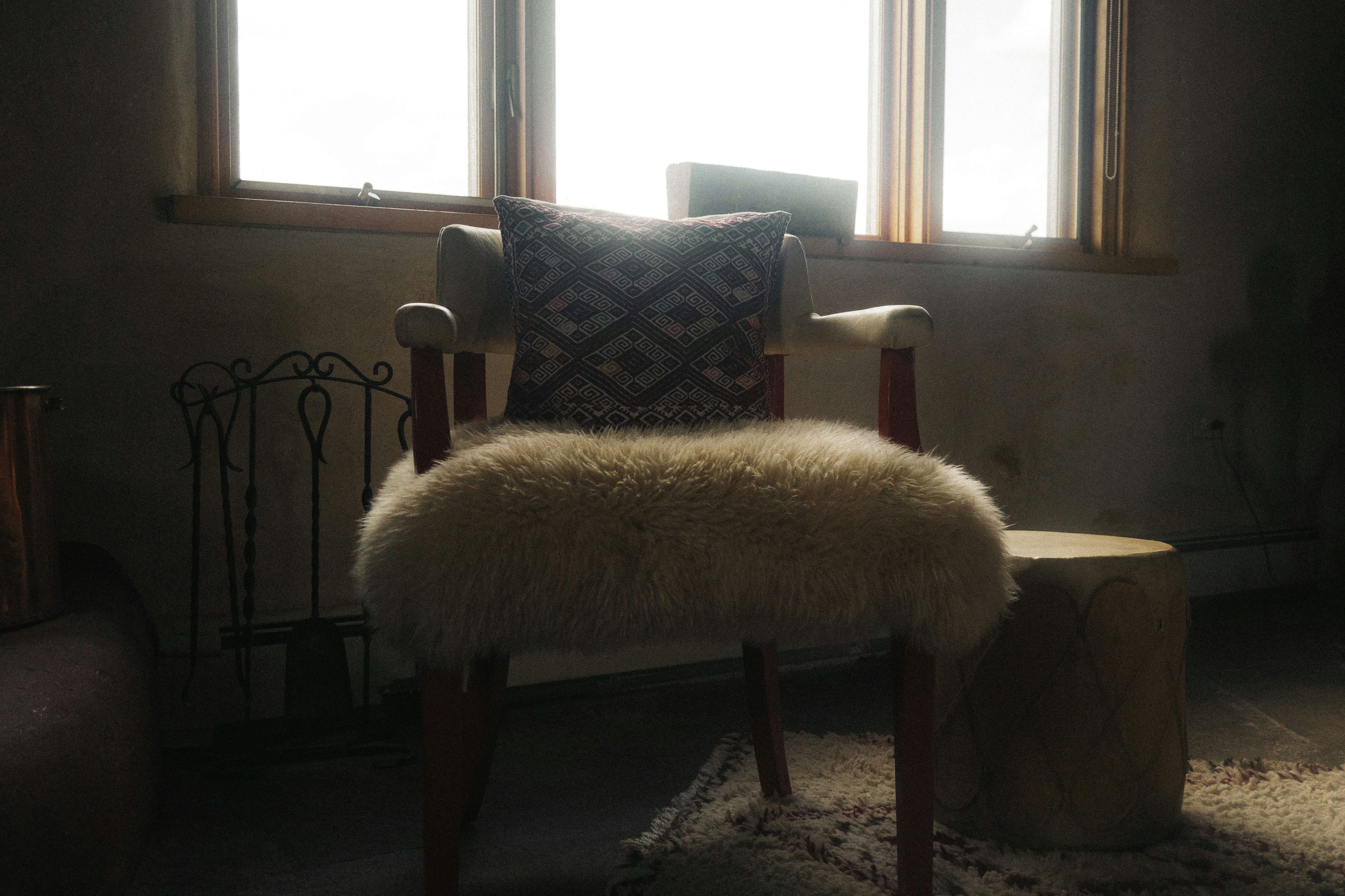 A rustic chair adorned with a fluffy throw and a patterned pillow, positioned near windows that softly illuminate the room's warm tones.