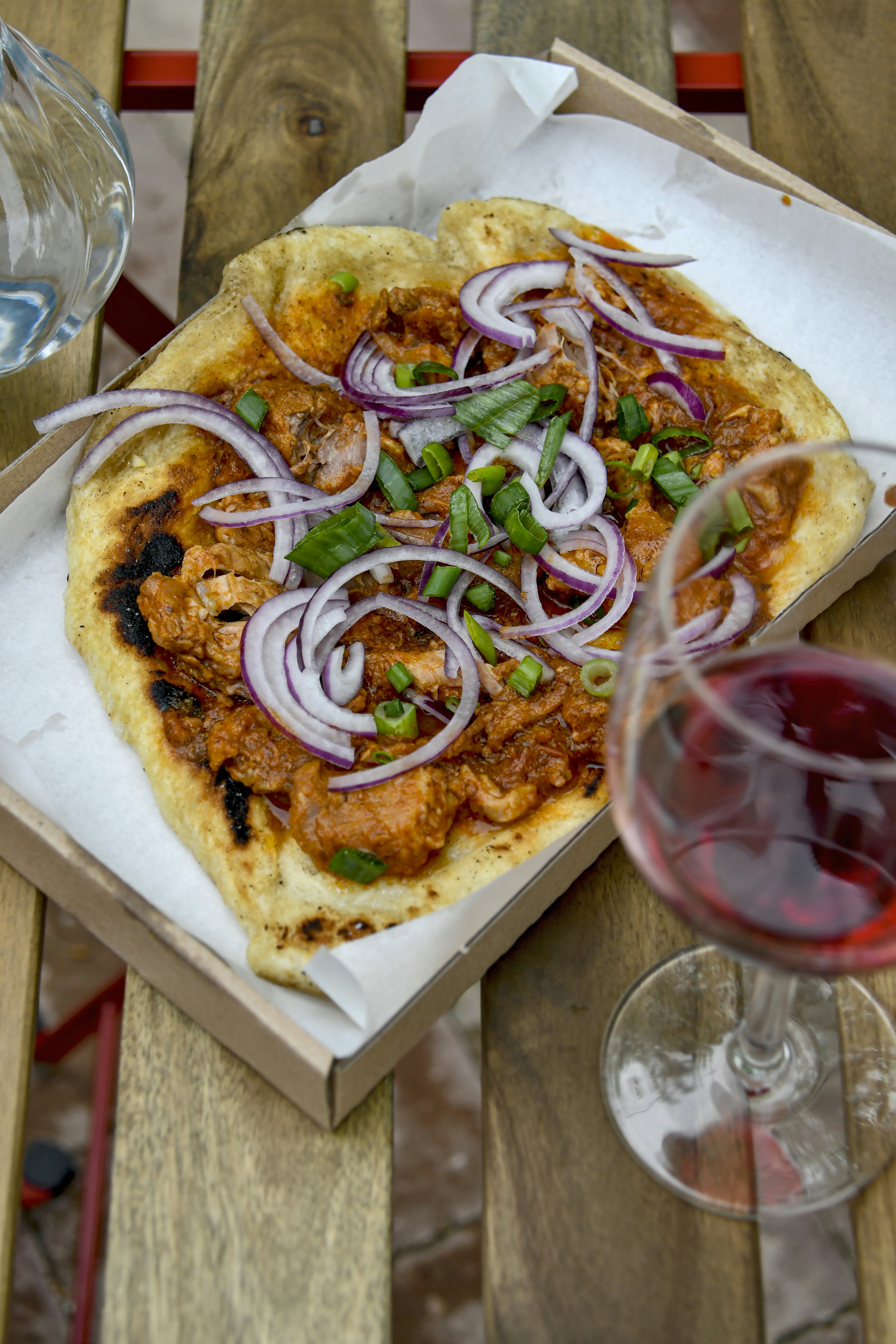 Flatbread-style pizza topped with red onions and chopped herbs sits in an open box on a wooden table, with a glass of red wine nearby.