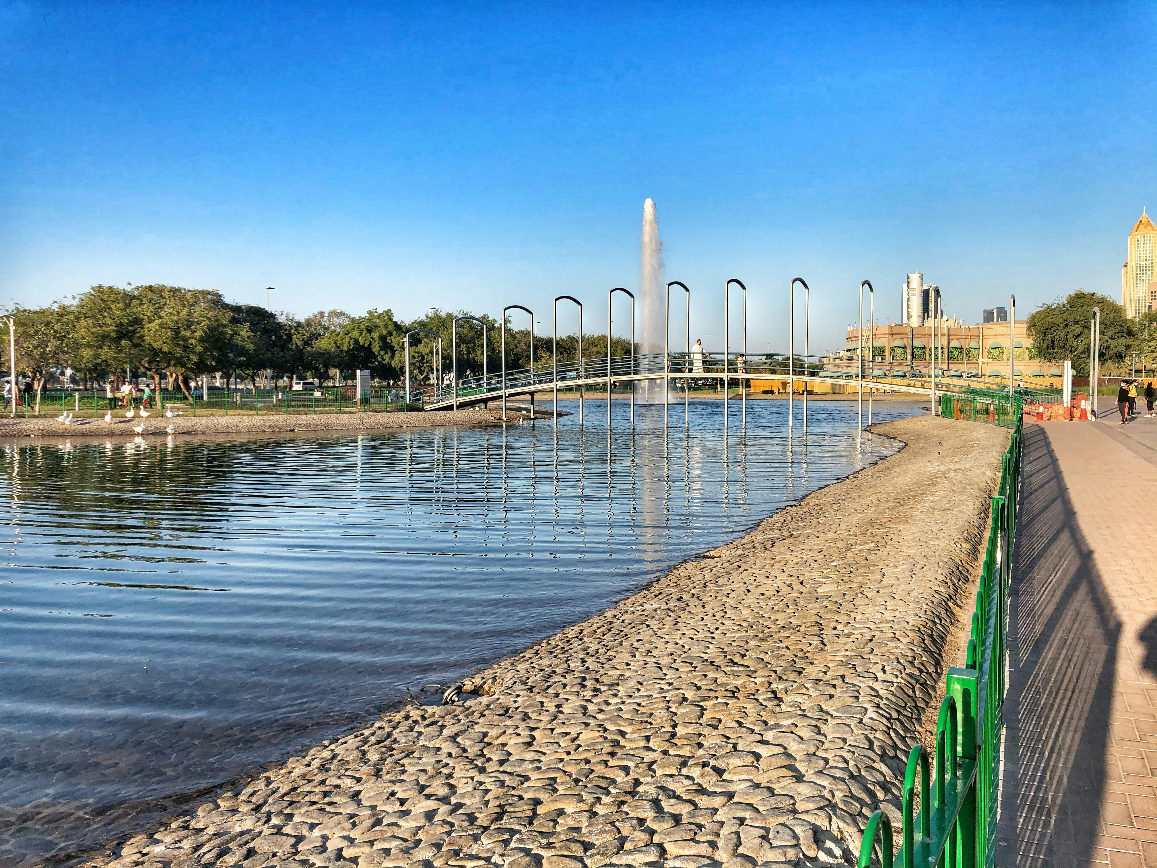 Serene lake with a fountain and footbridge in a city park under a clear blue sky.