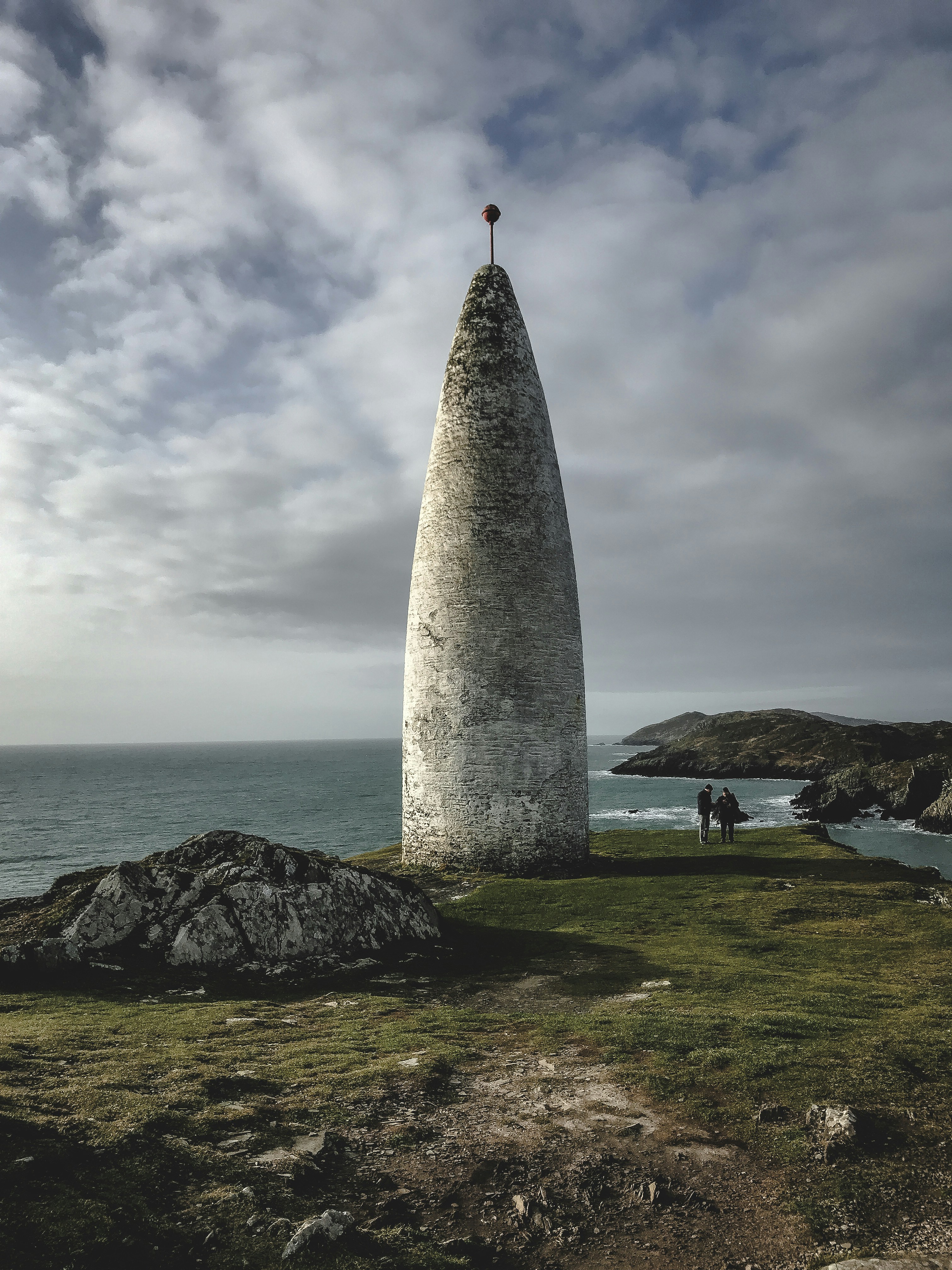 A towering white obelisk stands guard over the rugged coastline, with two figures exploring the landscape nearby. The dramatic sky adds depth to the scene.