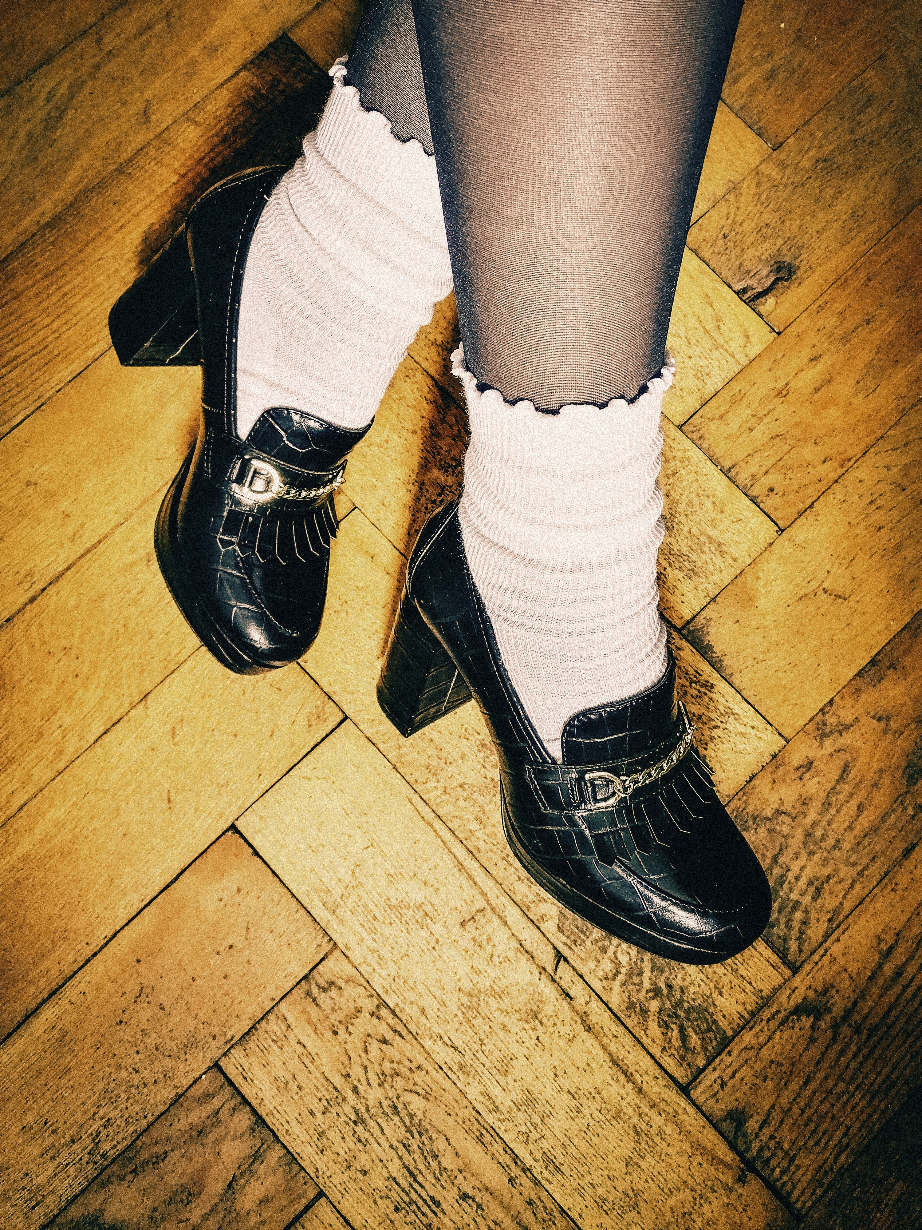Feet in white ribbed socks and black patent loafers on a honey-toned parquet, captured at an angle.