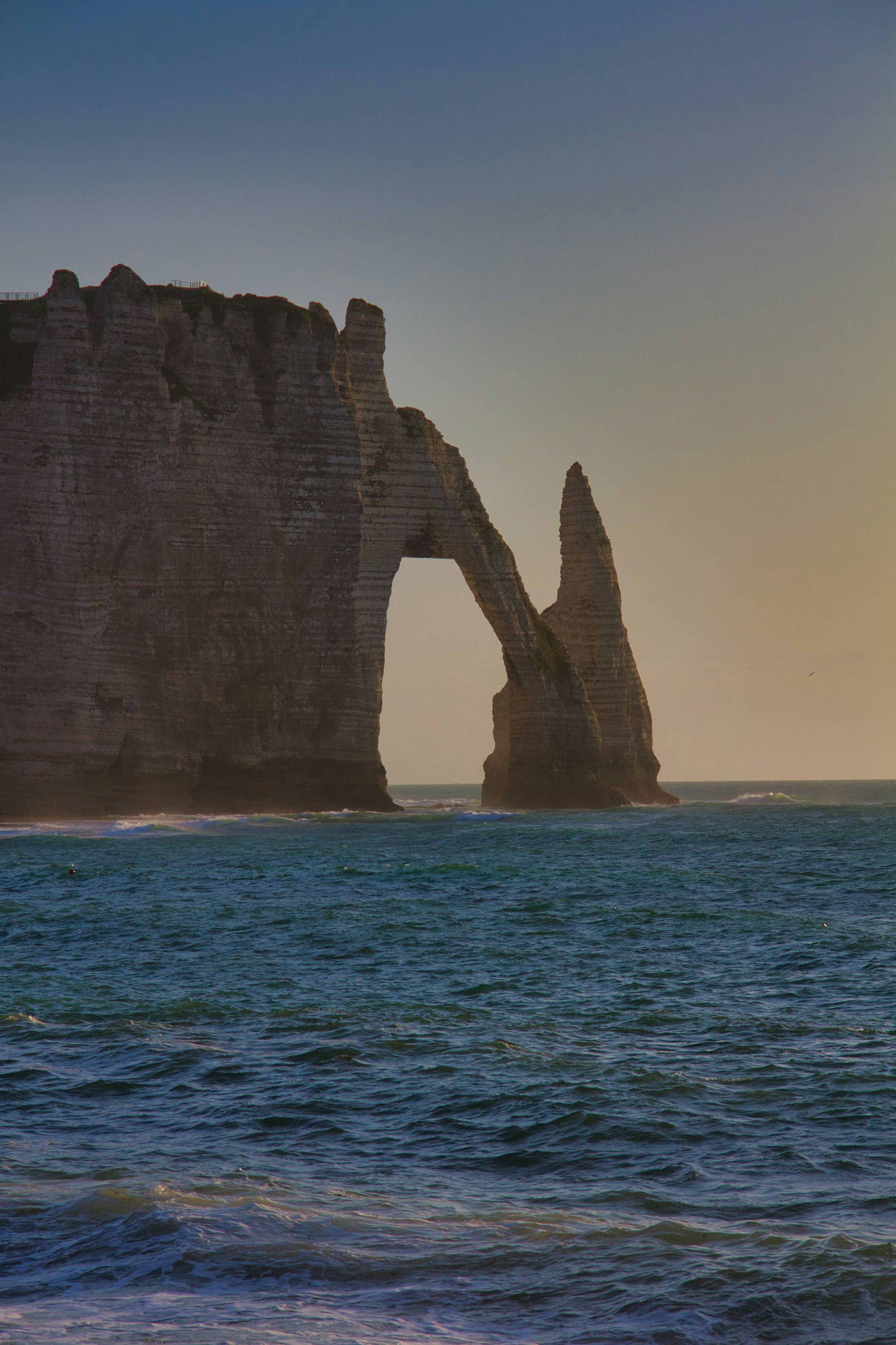 Majestic rock formation with a natural arch rising from the ocean, framed by a serene sky at dusk.