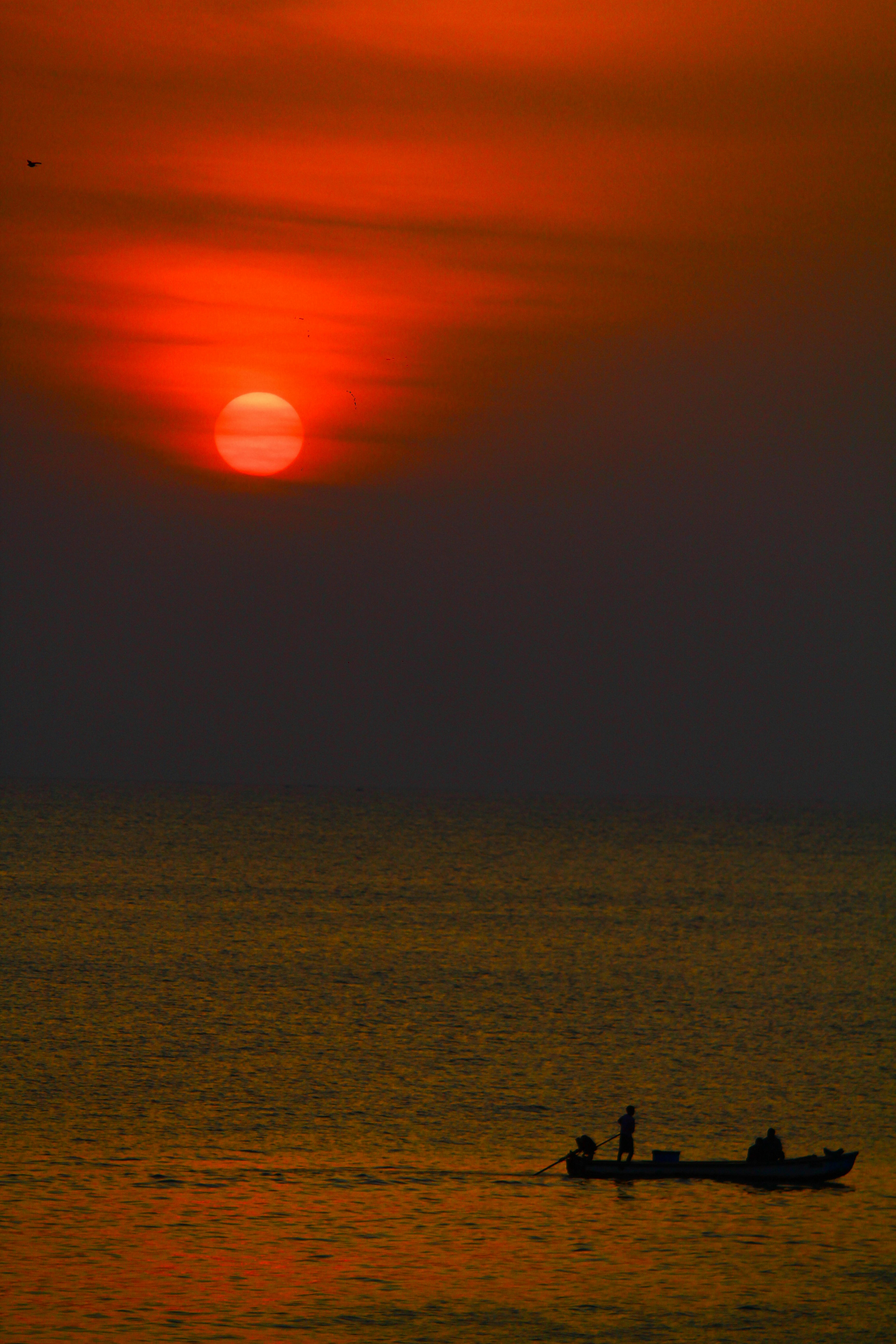 Silhouetted fishermen in a small boat glide across the shimmering sea as the sun sets, casting vibrant hues across the sky.