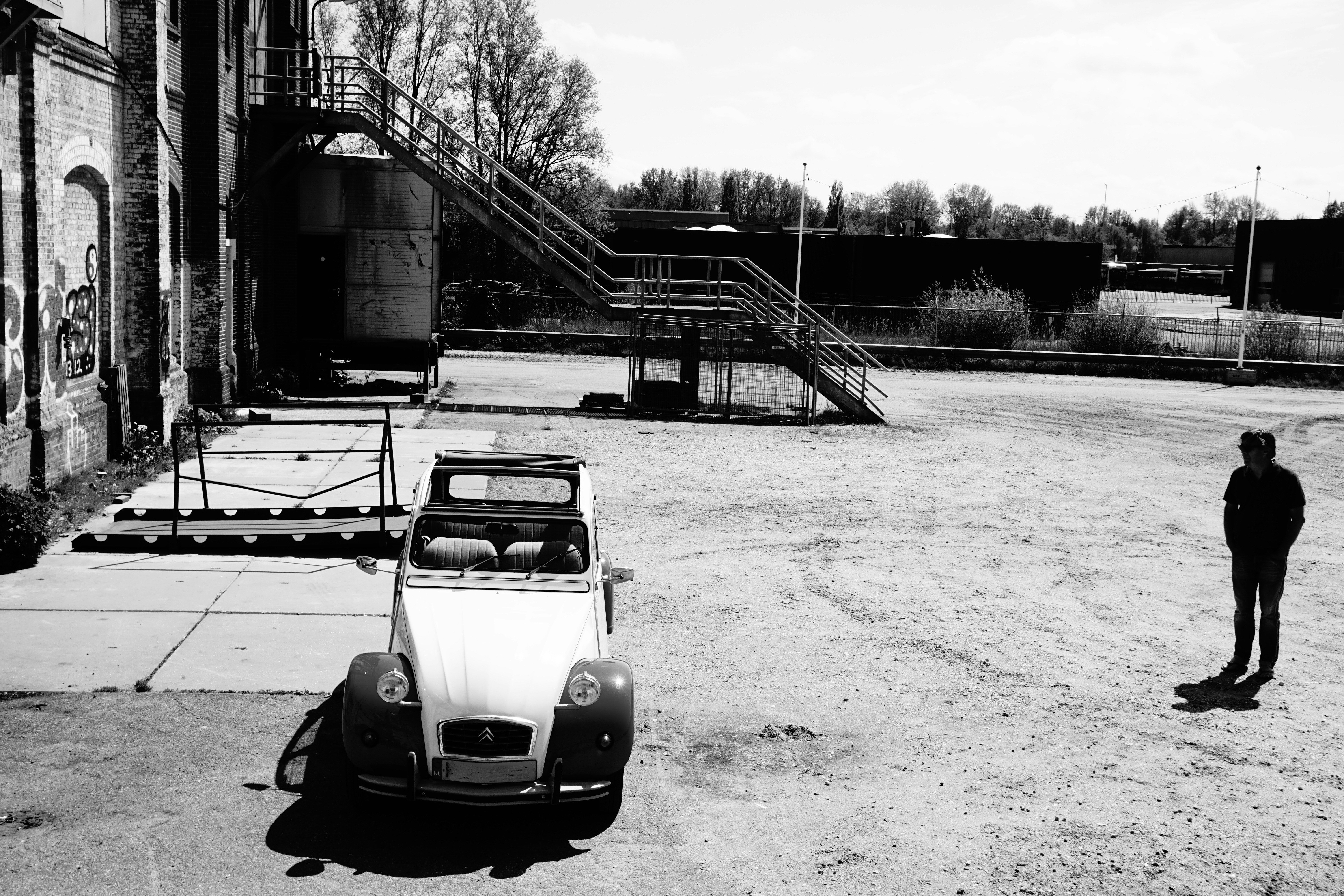 A vintage car parked on a weathered concrete surface, with a solitary figure standing nearby in an abandoned industrial setting.