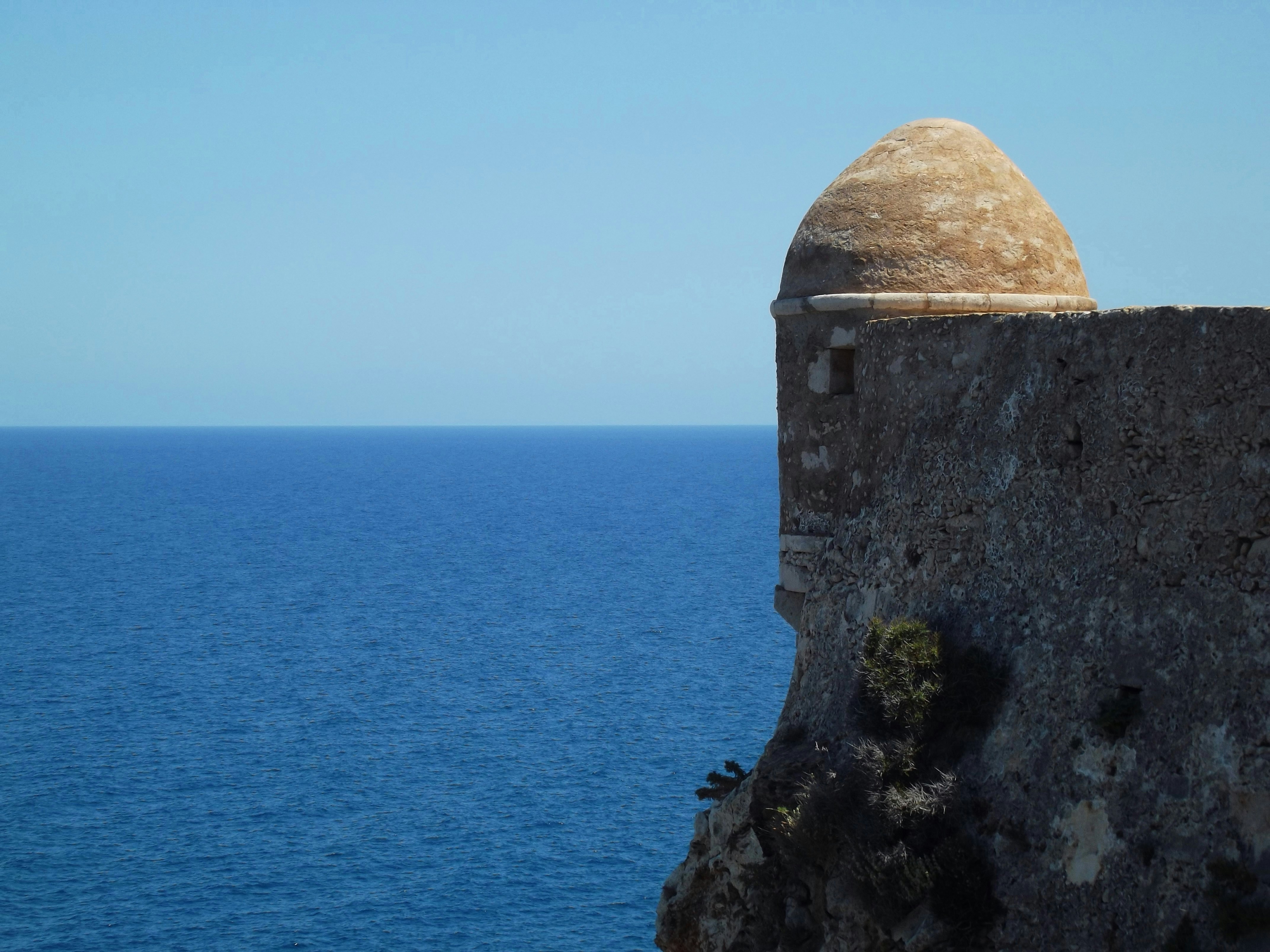 Stone fortress turret overlooking a vast, clear blue sea under a cloudless sky.