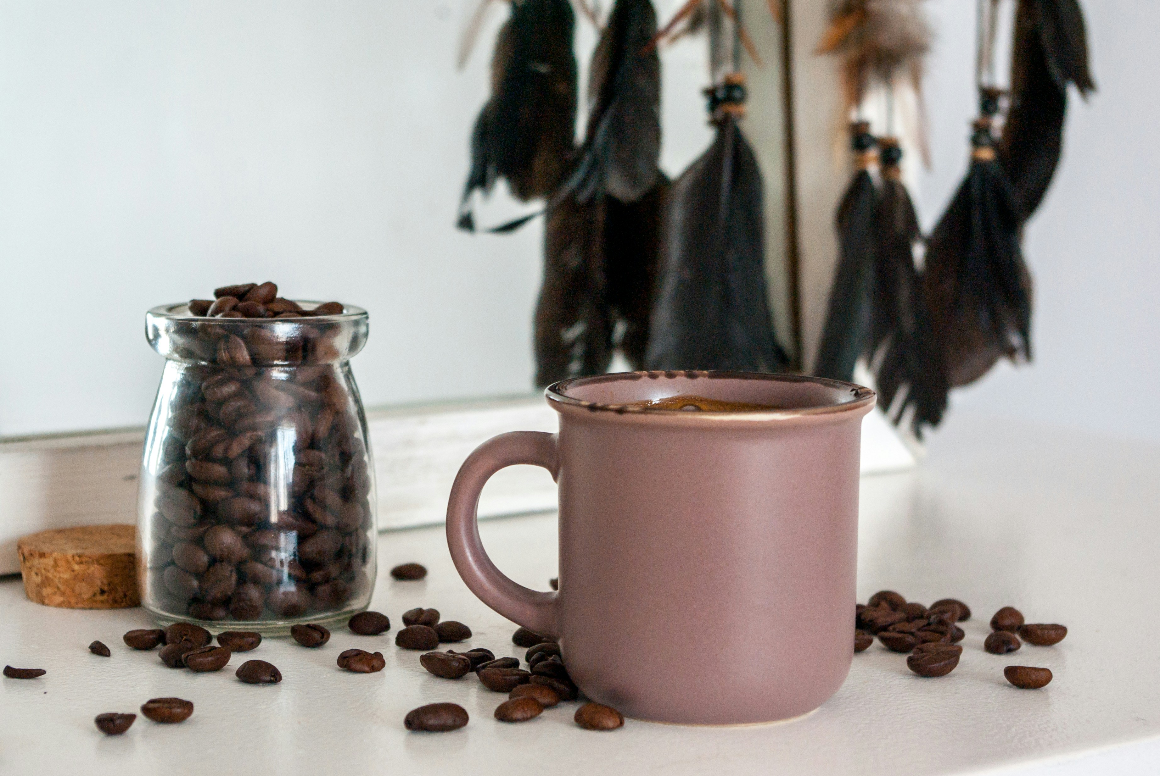 A cozy scene featuring a pink mug filled with coffee, surrounded by scattered coffee beans and a jar of whole beans, complemented by decorative feathers in the background.