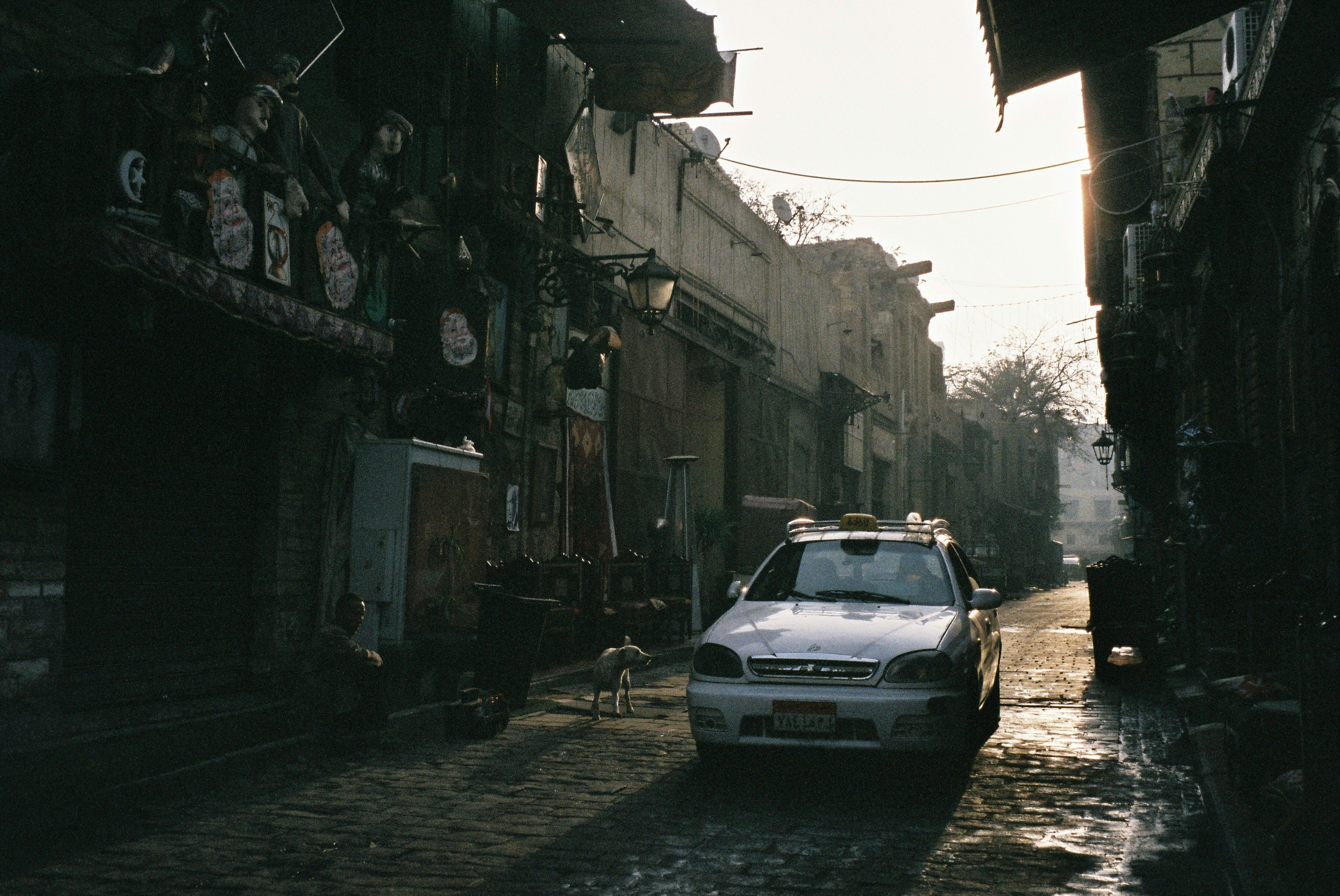Car driving through a narrow, sunlit alley flanked by weathered buildings.