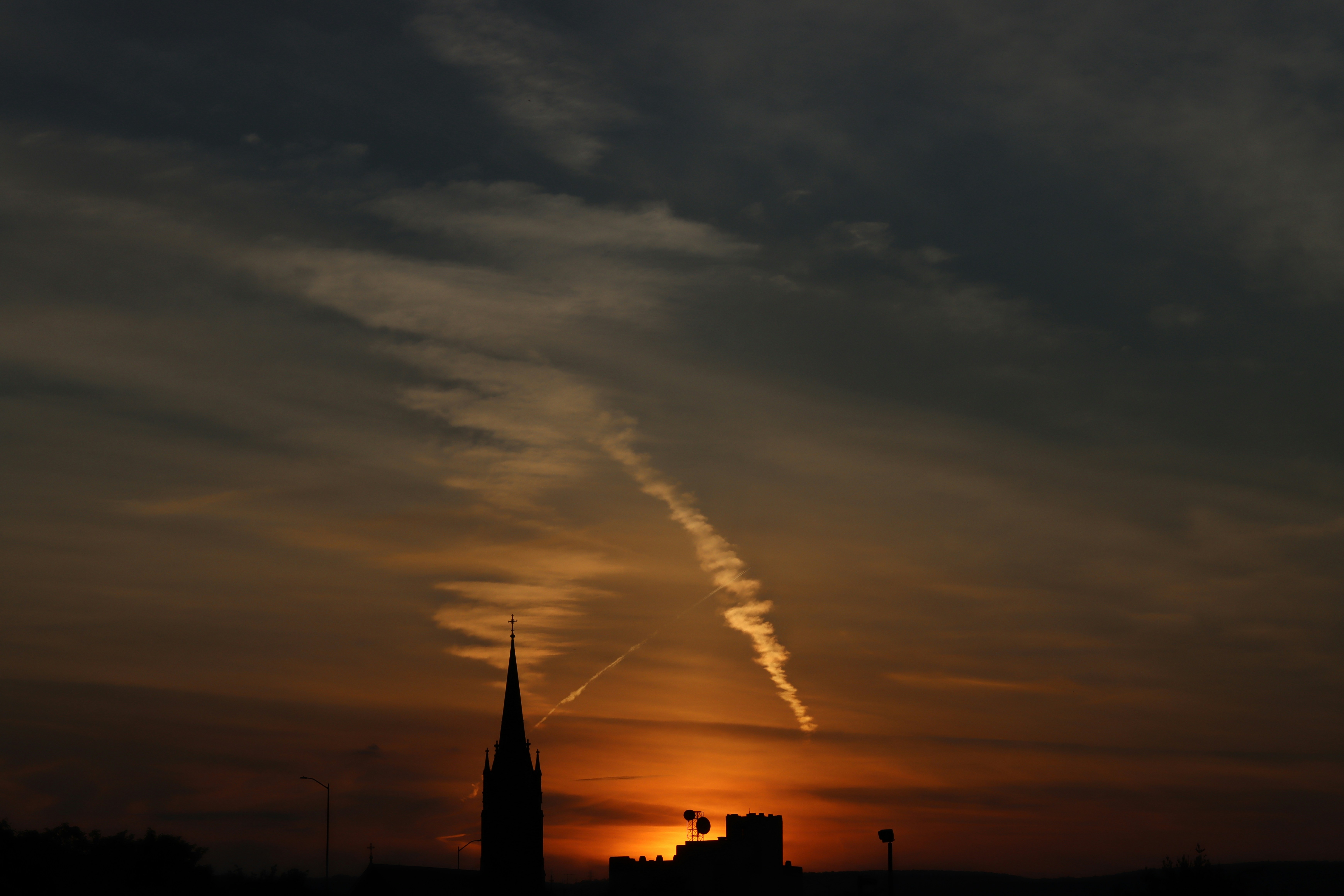 Silhouette of a city skyline at sunset with dramatic clouds and contrails.