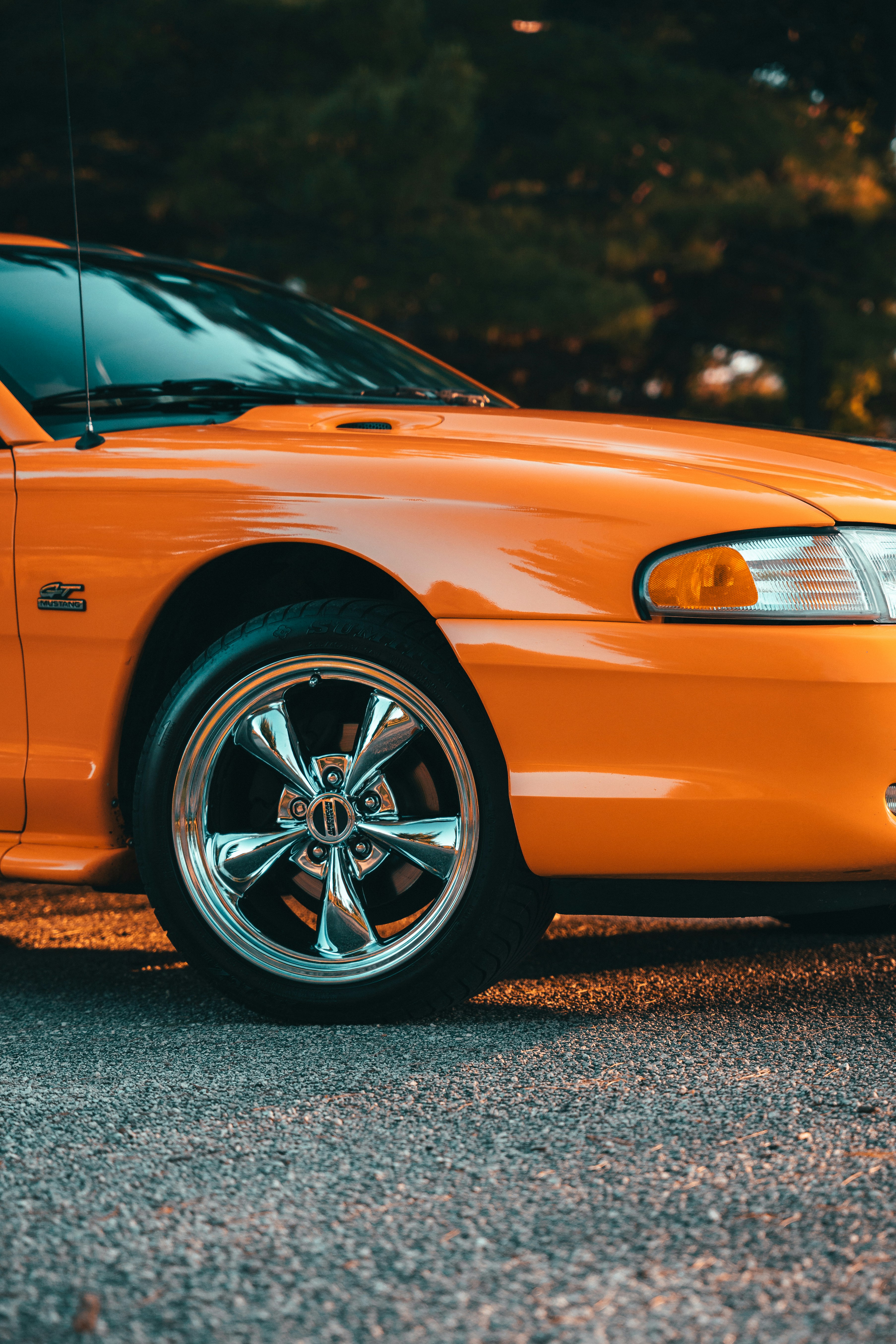 Close-up of a bright orange car showcasing its sleek design and polished chrome wheels against a blurred natural backdrop.