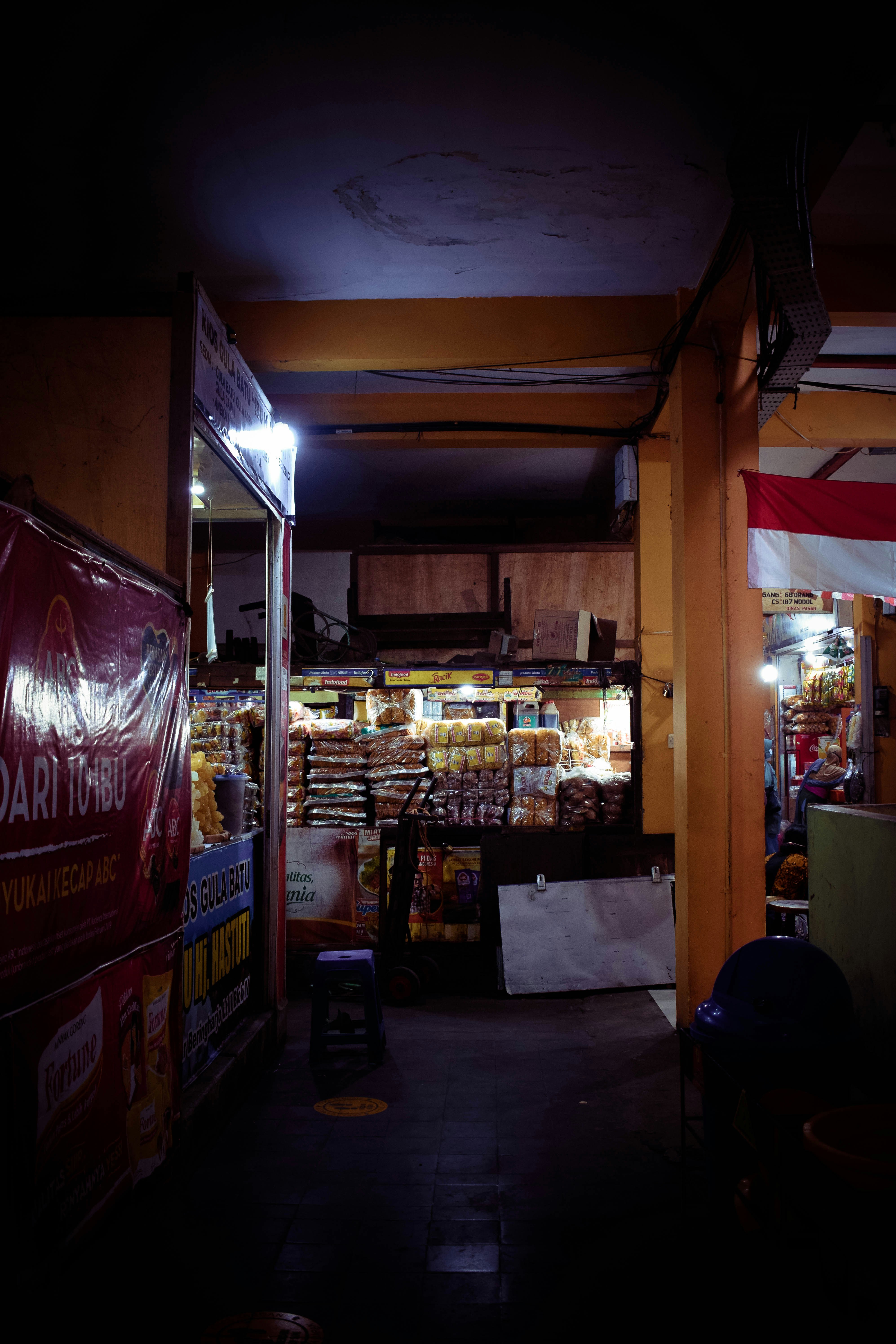Dimly lit alleyway of a bustling night market, showcasing shelves filled with snacks and goods under warm lights. A flag hangs in the background.