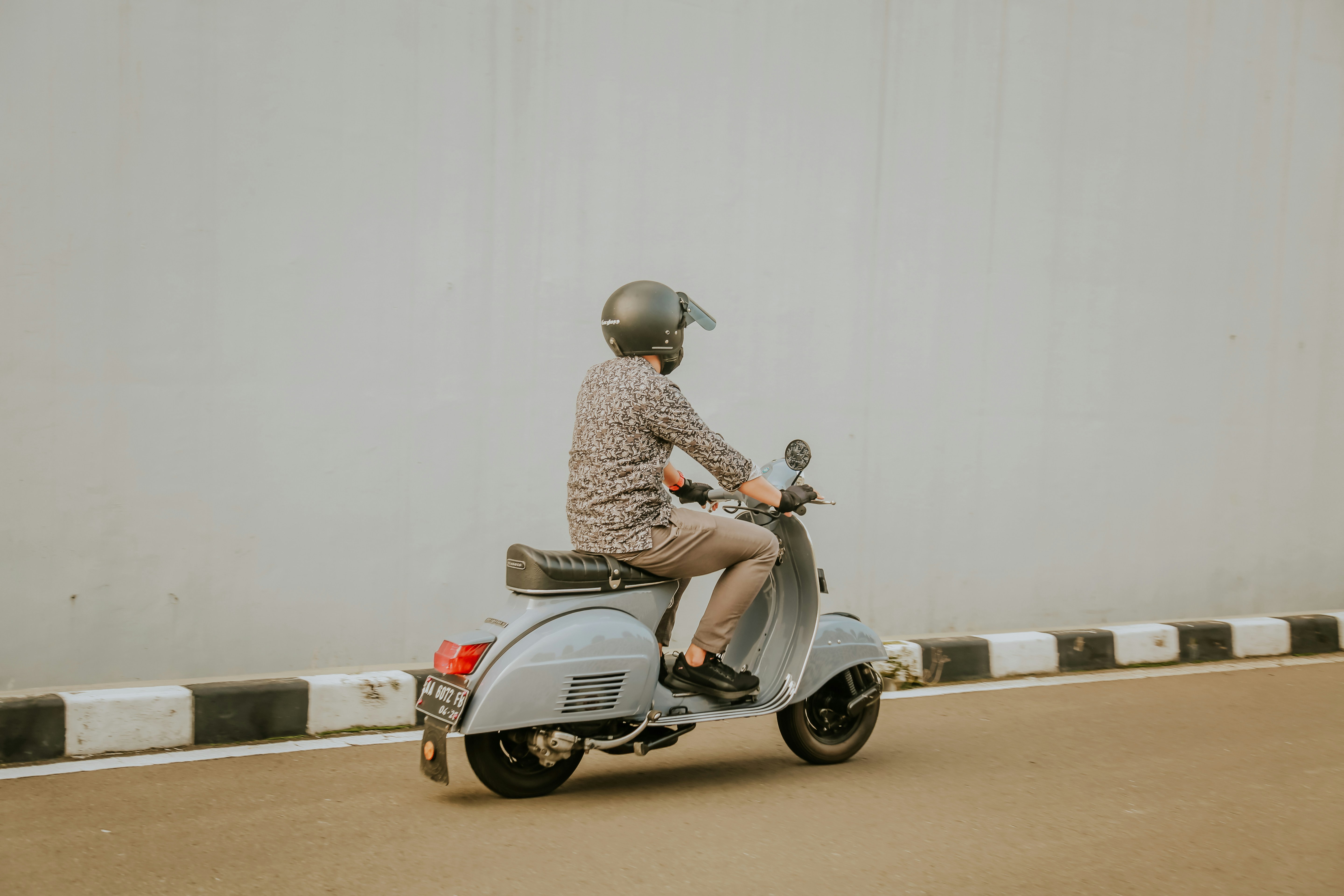 A person riding a vintage scooter along a smooth road, wearing a helmet and stylish attire, with a minimalist background.