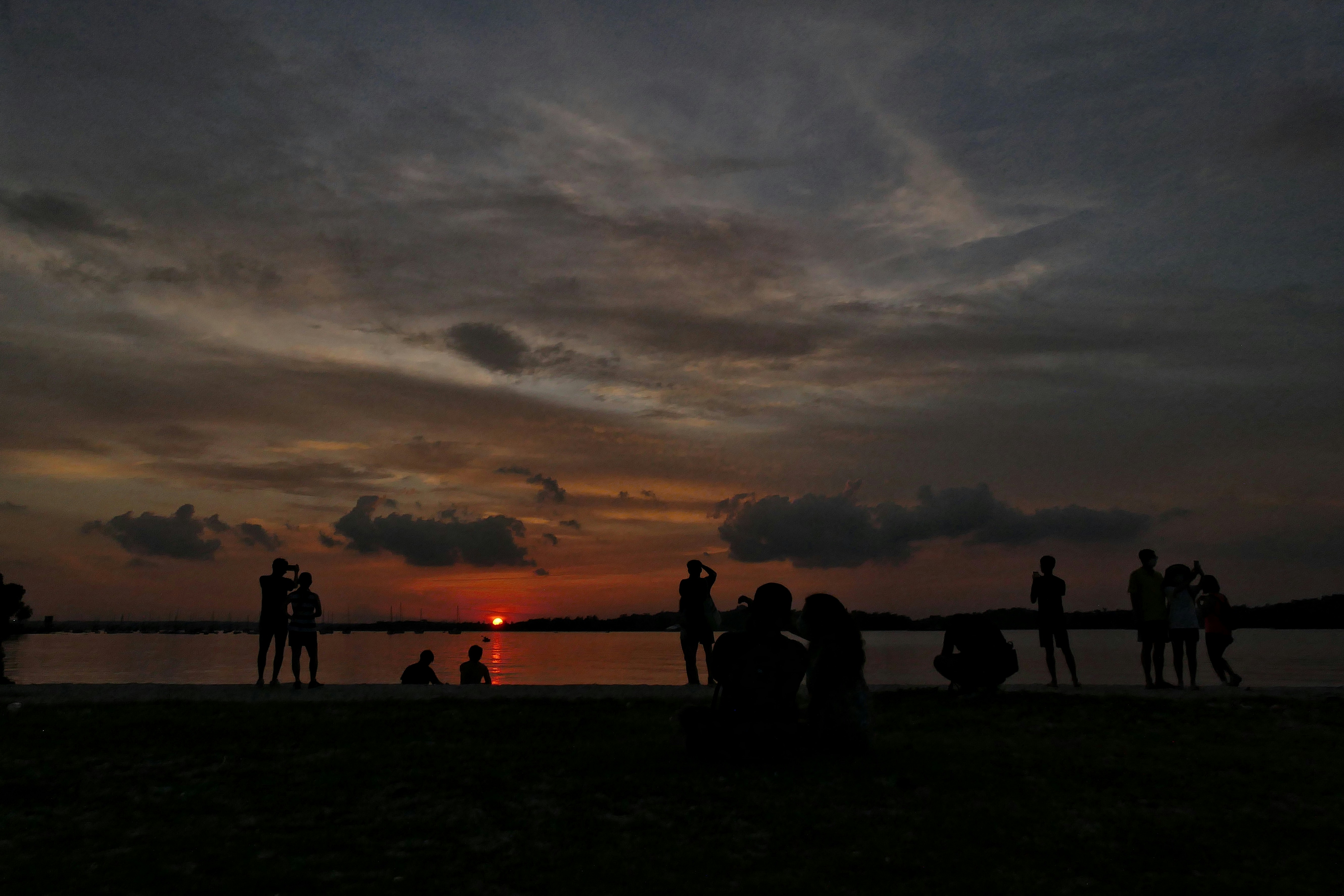 Groups of people stand and sit along a darkened shoreline as a low sun sinks over a calm sea, casting warm orange light across the clouds. Silhouettes contrast with the fiery sky, capturing a serene beach moment at dusk.