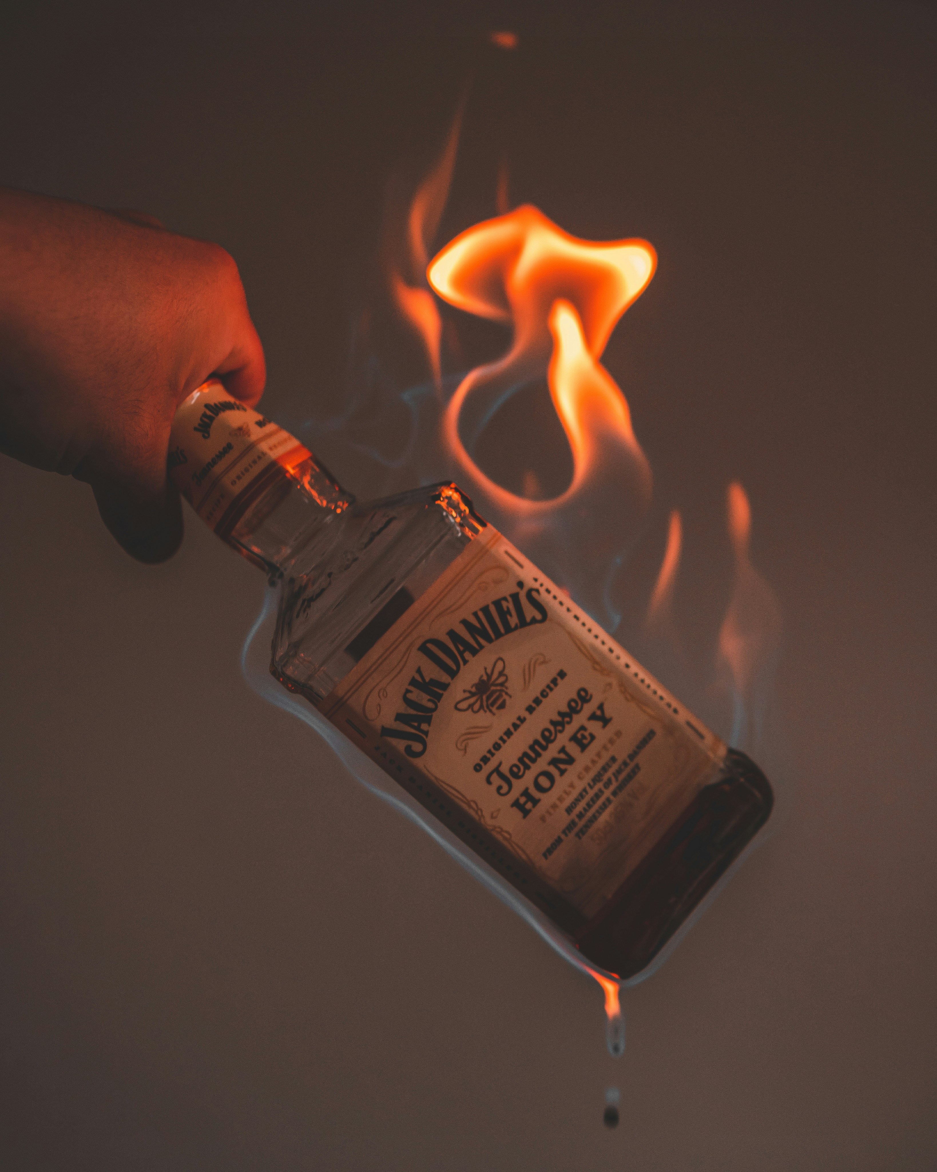 A hand holds a bottle of Jack Daniel's Tennessee Honey, engulfed in flames with honey dripping from the bottom.