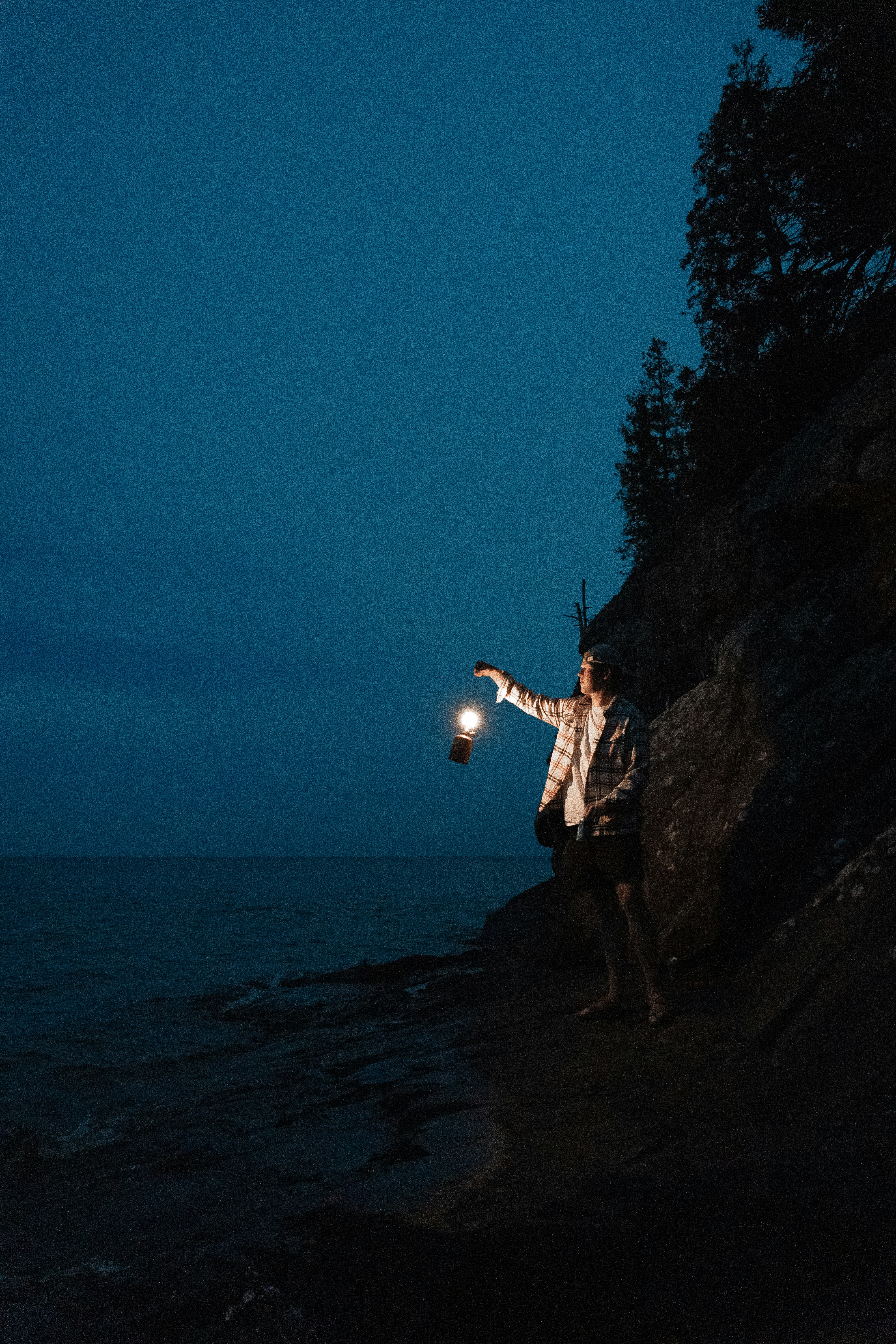 A figure stands on a rocky shoreline, holding a lantern aloft against the twilight sky. The calm waters reflect the soft glow of the light.