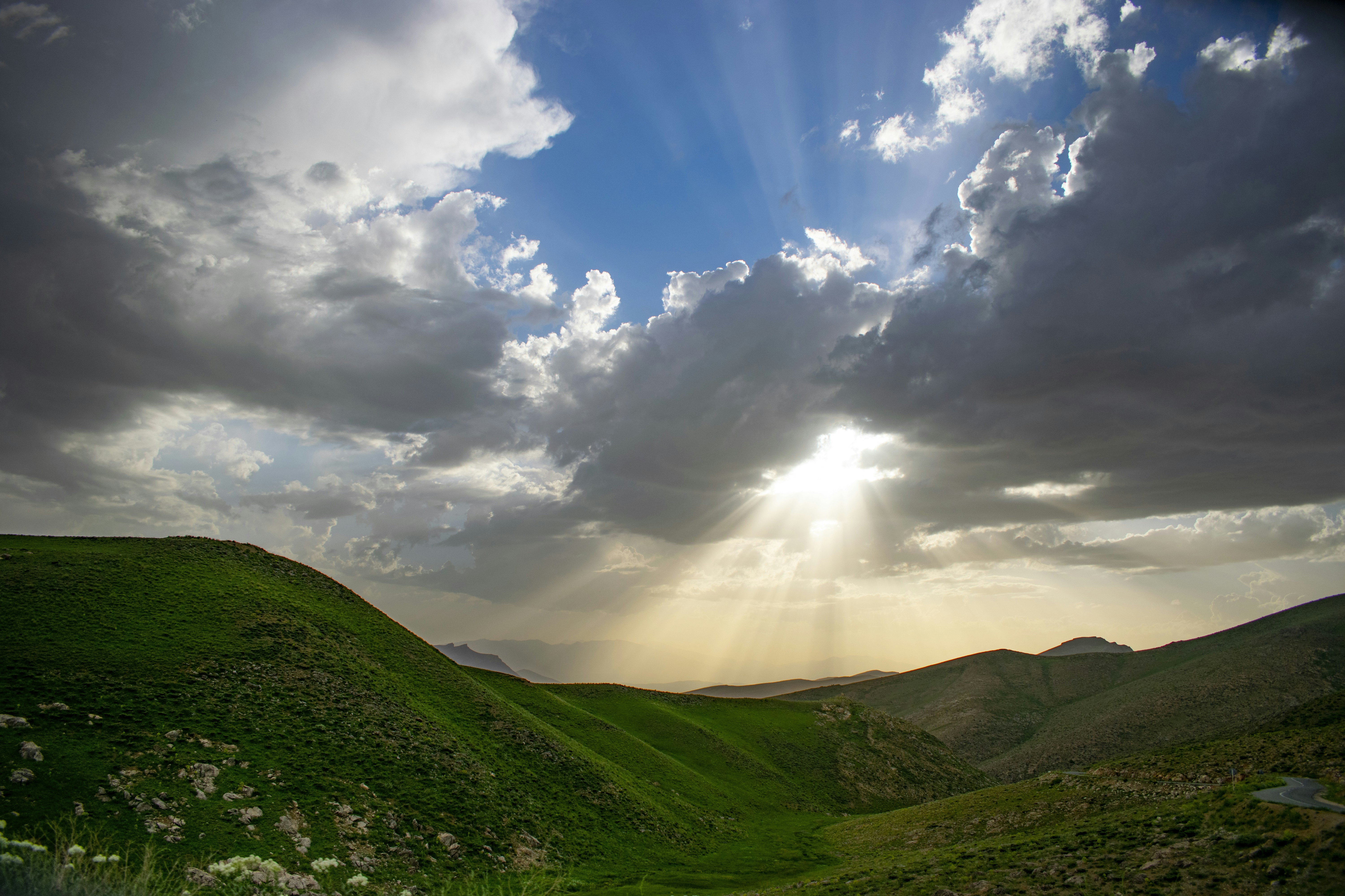 Sunlight breaks through dramatic clouds over rolling green hills, creating a serene landscape. The scene captures the essence of nature's tranquility.