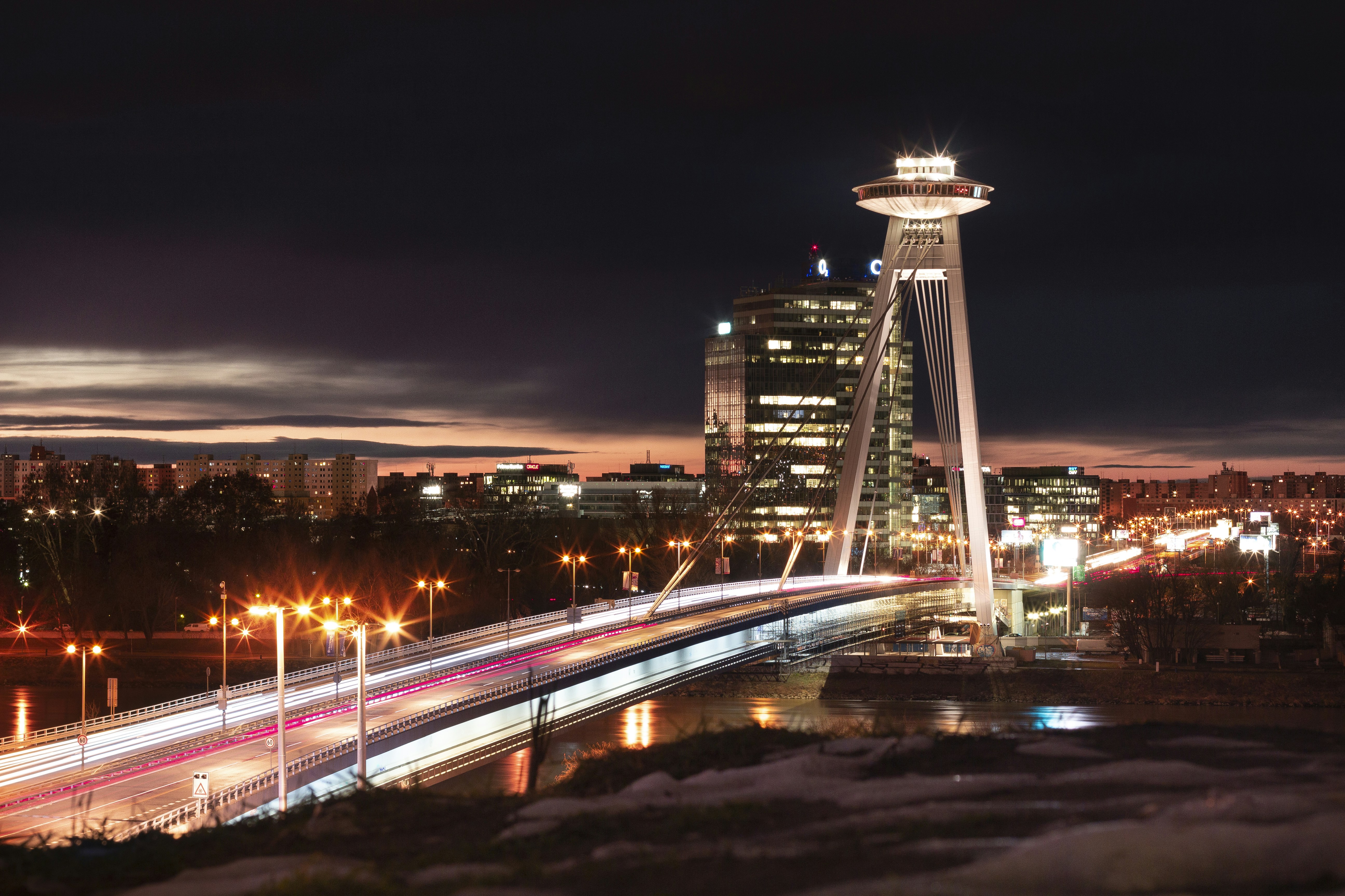 A modern bridge illuminated against a twilight sky, showcasing vibrant light trails from passing vehicles. The scene captures the dynamic interplay between architecture and motion.