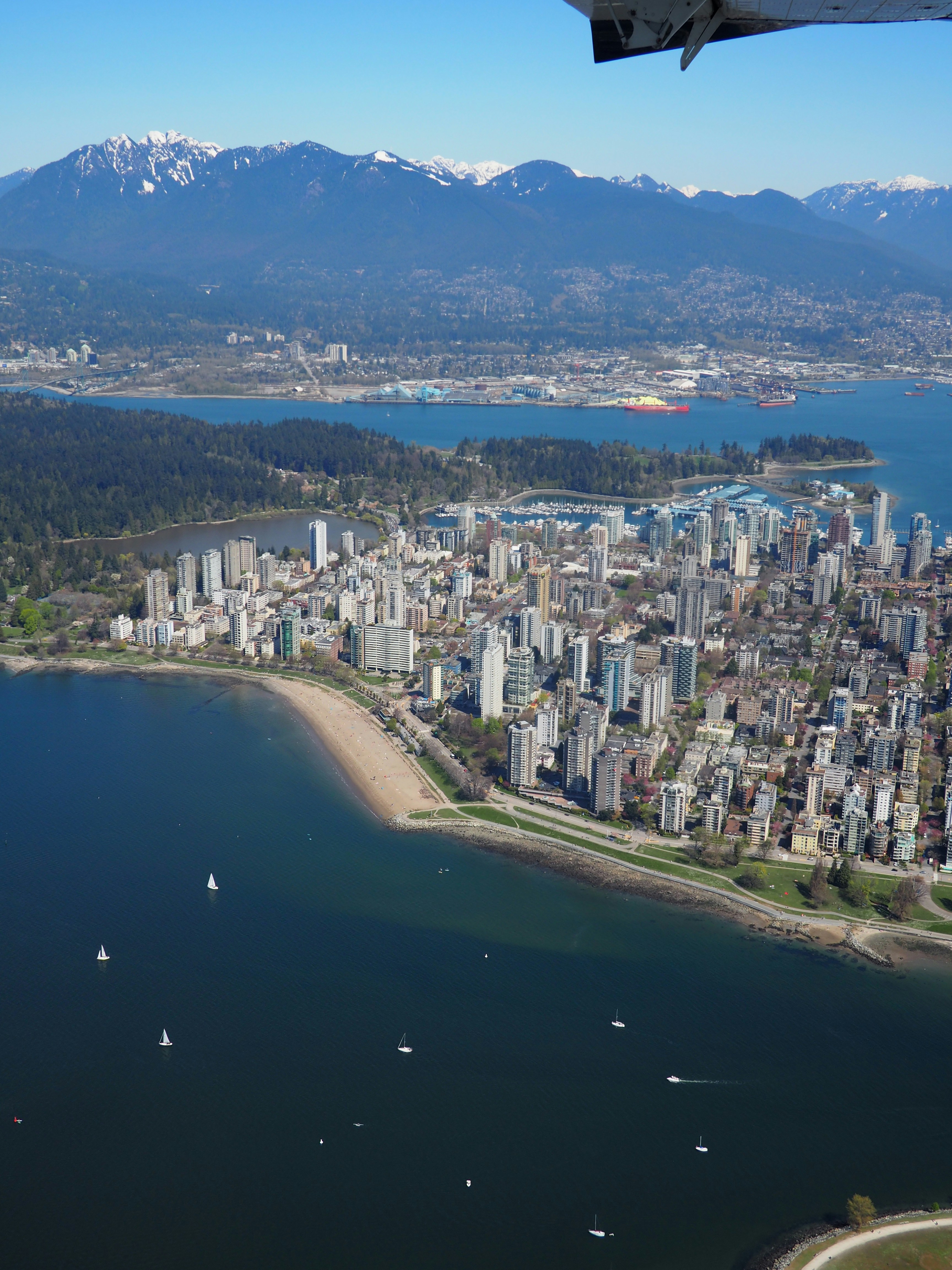 Aerial view of a vibrant coastal city with skyscrapers and lush greenery, framed by mountains and a shimmering bay. Sailboats dot the water's surface.