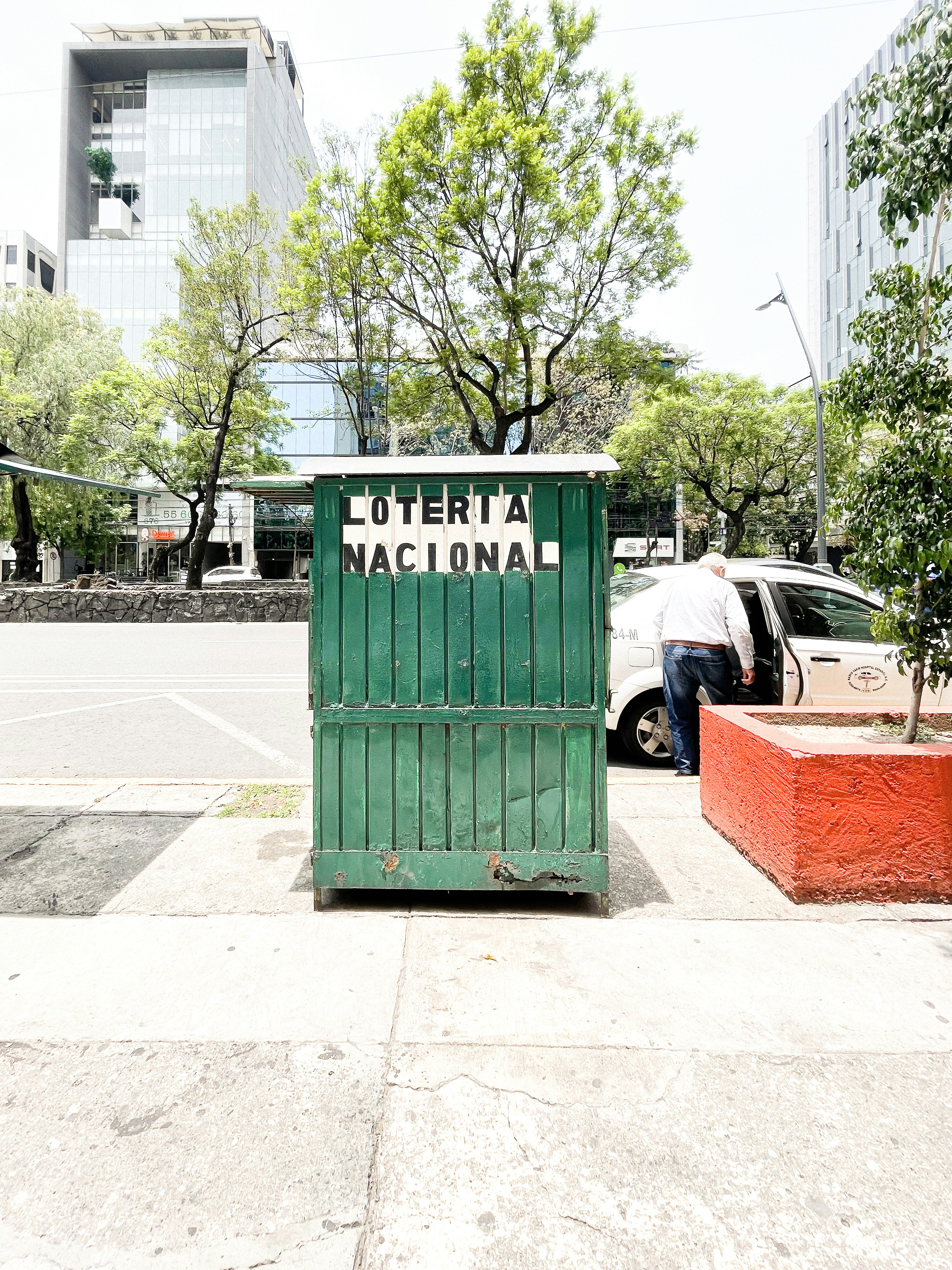 A green lottery booth stands prominently on a city sidewalk, surrounded by urban greenery and modern architecture. The booth features bold lettering reading 'LOTERÍA NACIONAL.'