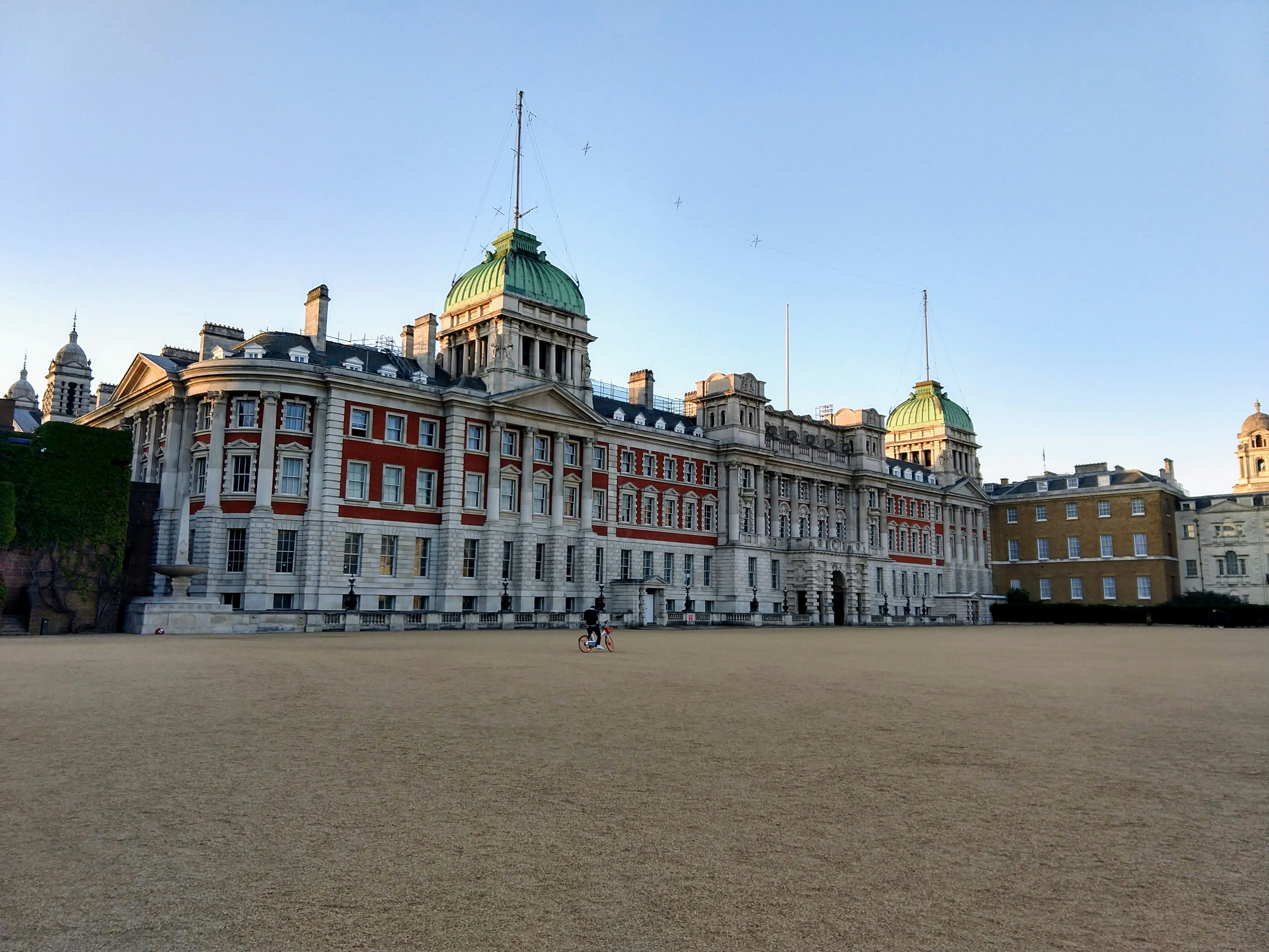 Wide-angle photograph of a grand neoclassical building with verdigris domes along a broad gravel foreground under a clear blue sky. The composition emphasizes architectural symmetry and historic grandeur.