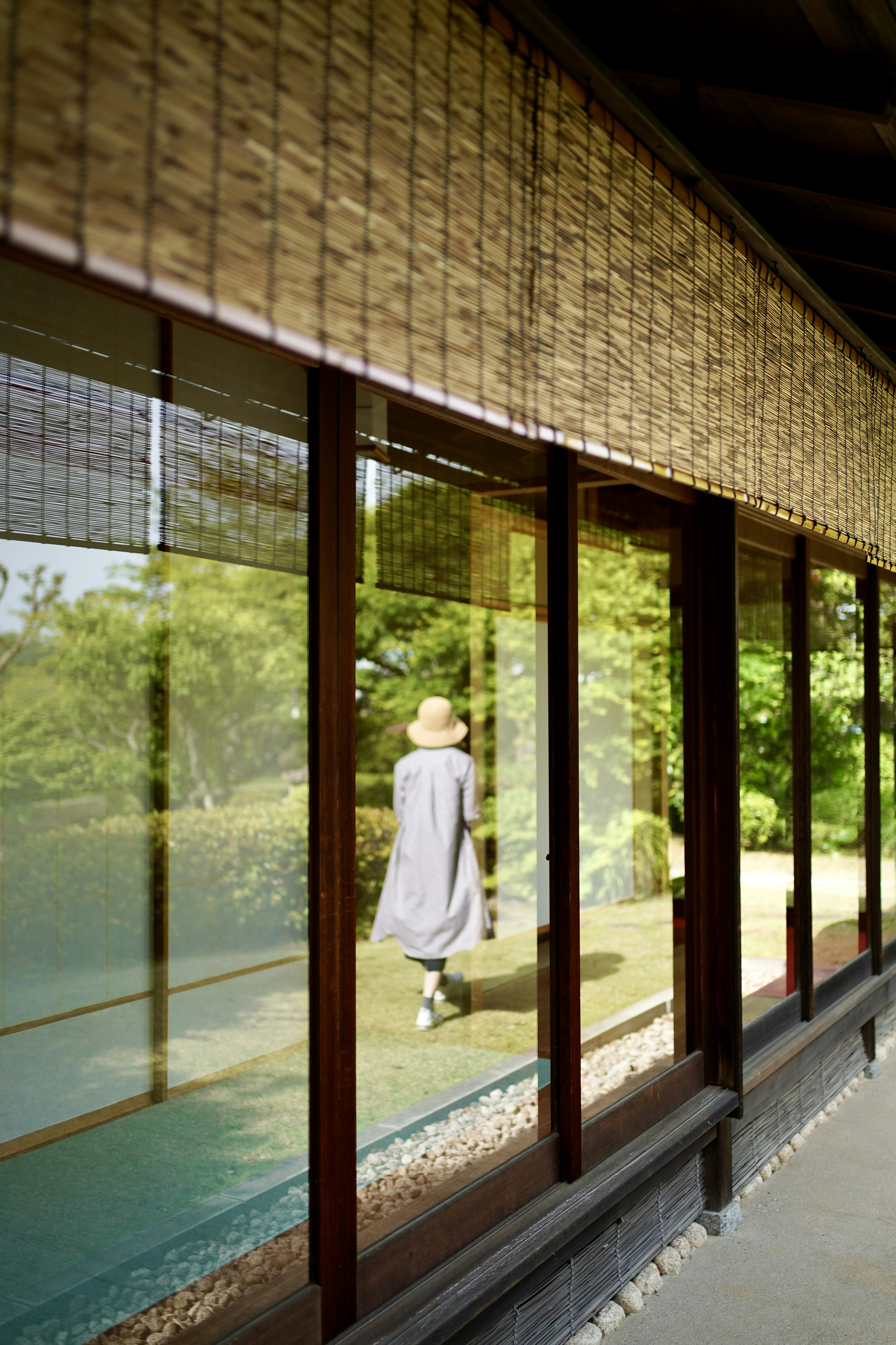 A woman in a straw hat and dress is reflected in the glass of a tea room in a Japanese garden. | woman in white dress standing near glass window