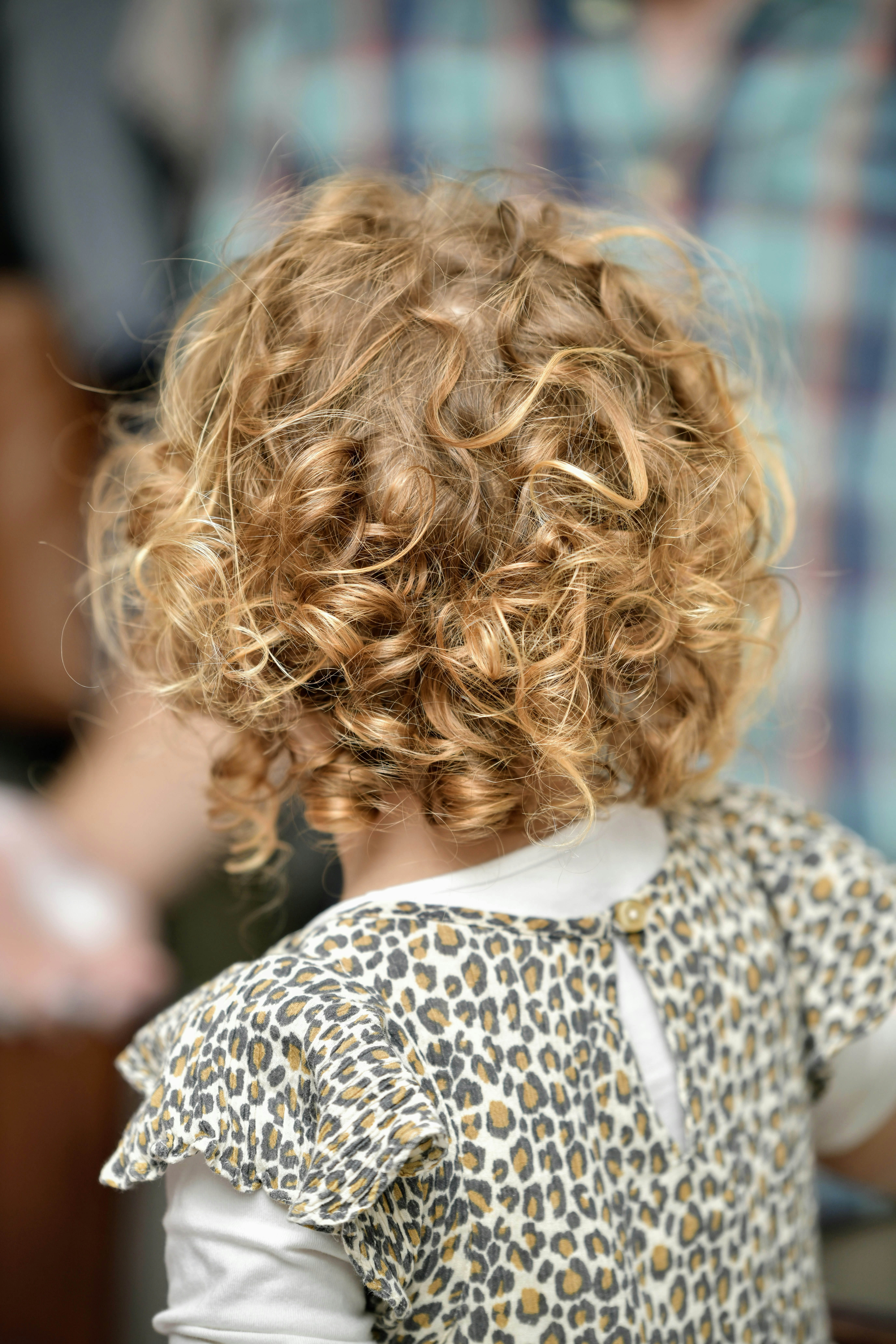 Curly-haired child in a leopard-print outfit, seen from behind, exploring a lively environment.