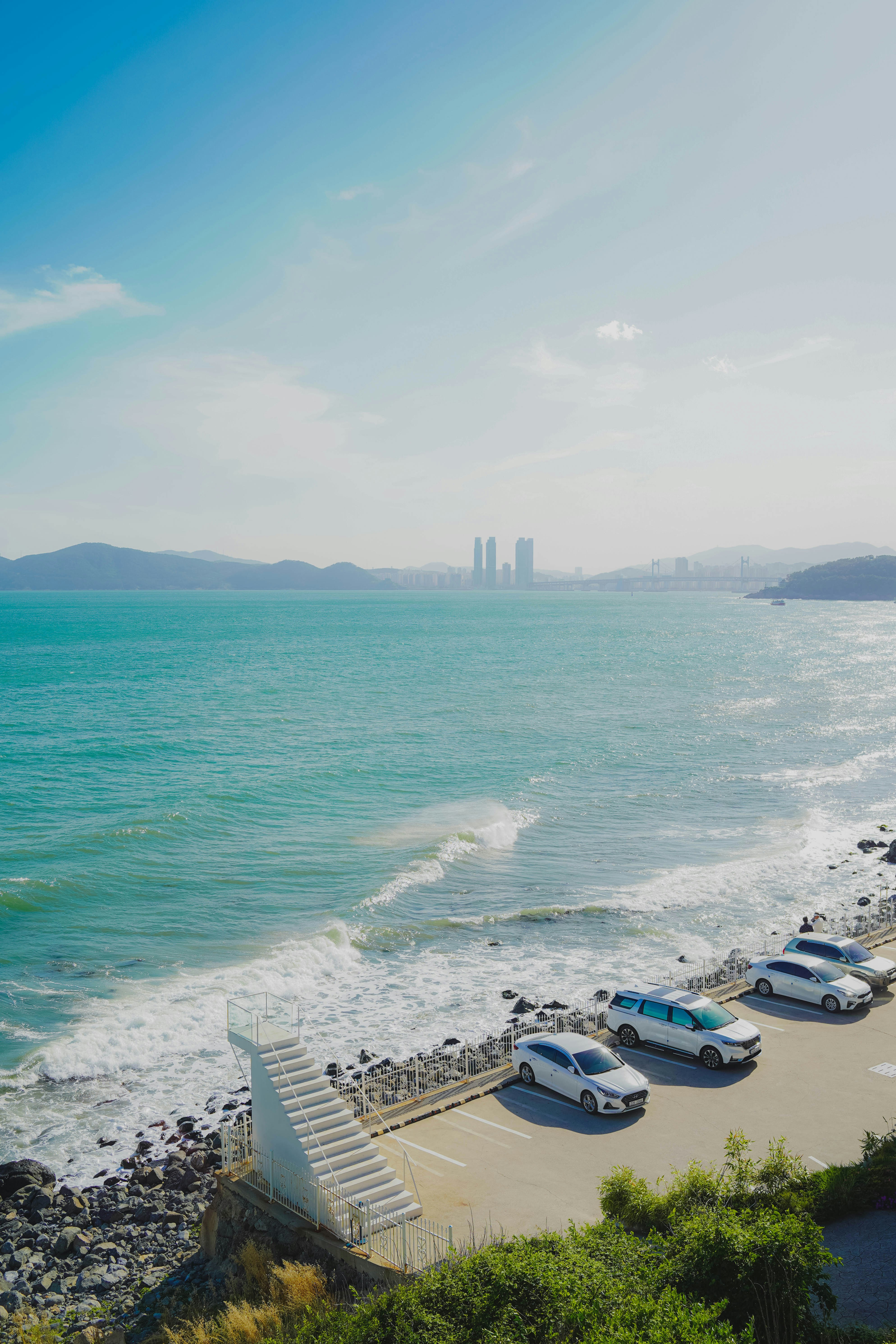 A serene coastal scene featuring parked cars along a rocky shore, with waves lapping at the coastline and distant city skyscrapers rising against a clear sky.