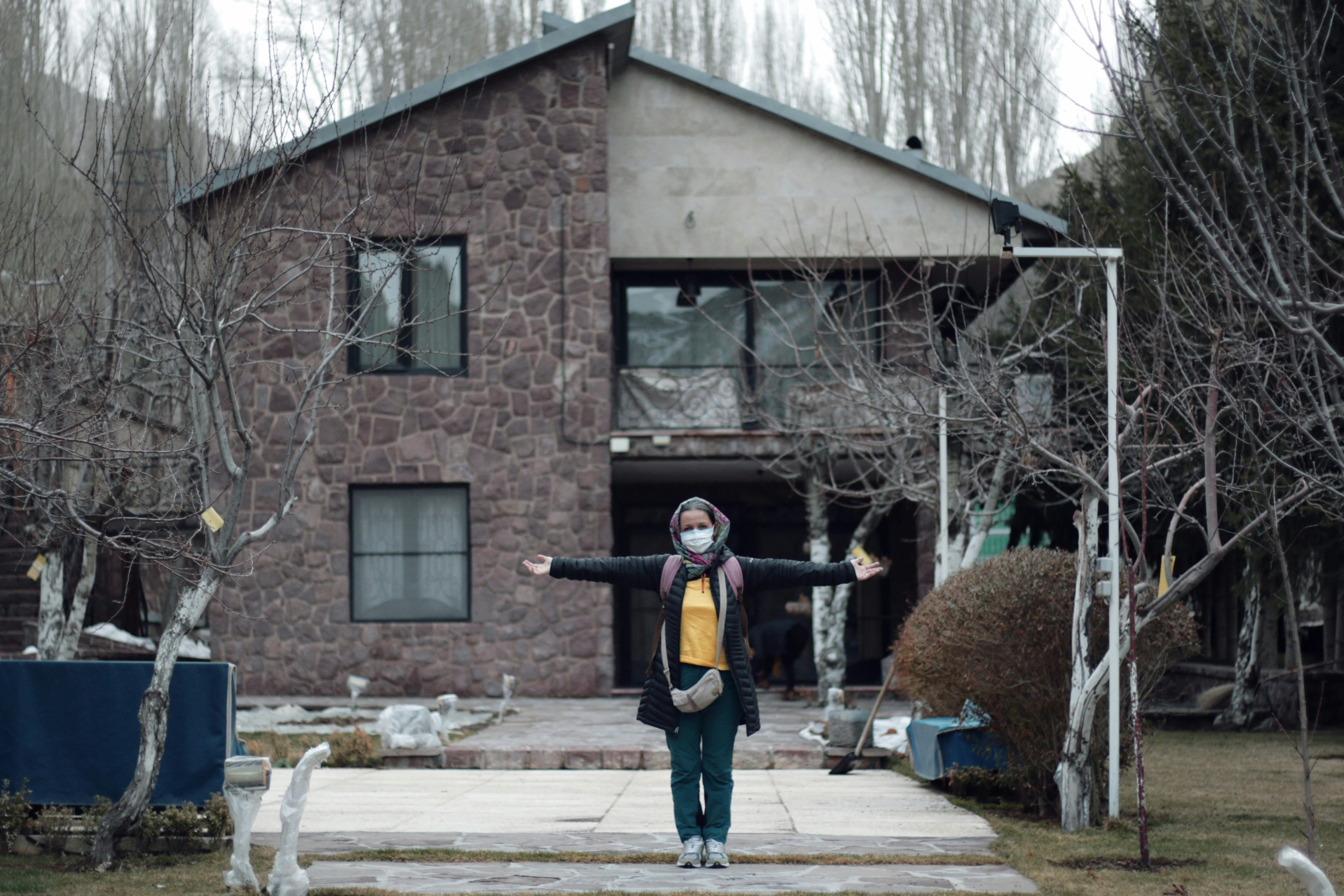 A woman stands with arms outstretched in front of a rustic stone house, surrounded by barren trees and a tranquil garden.