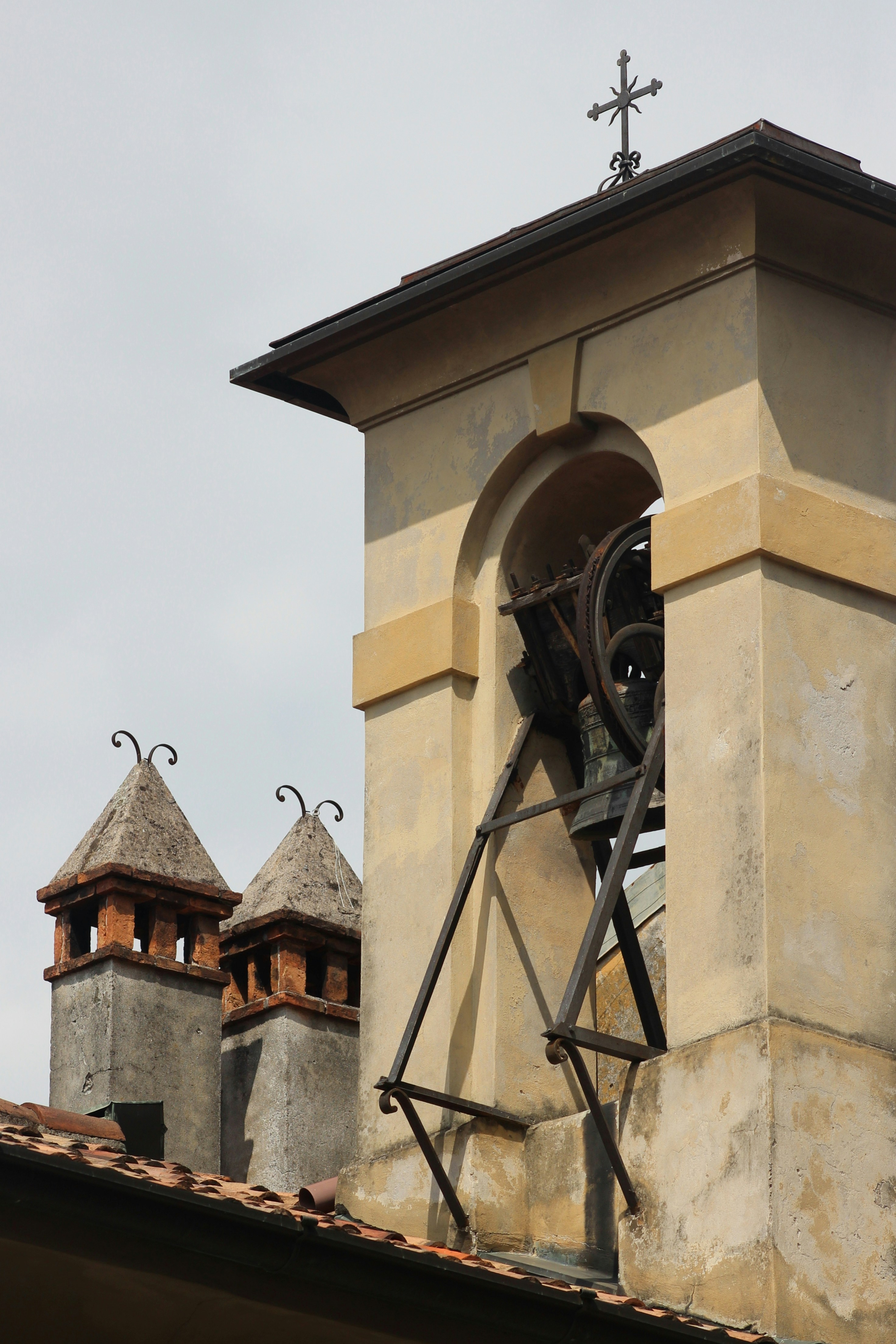 Weathered bell tower adorned with a cross, featuring intricate mechanical elements and surrounded by rustic rooftops.