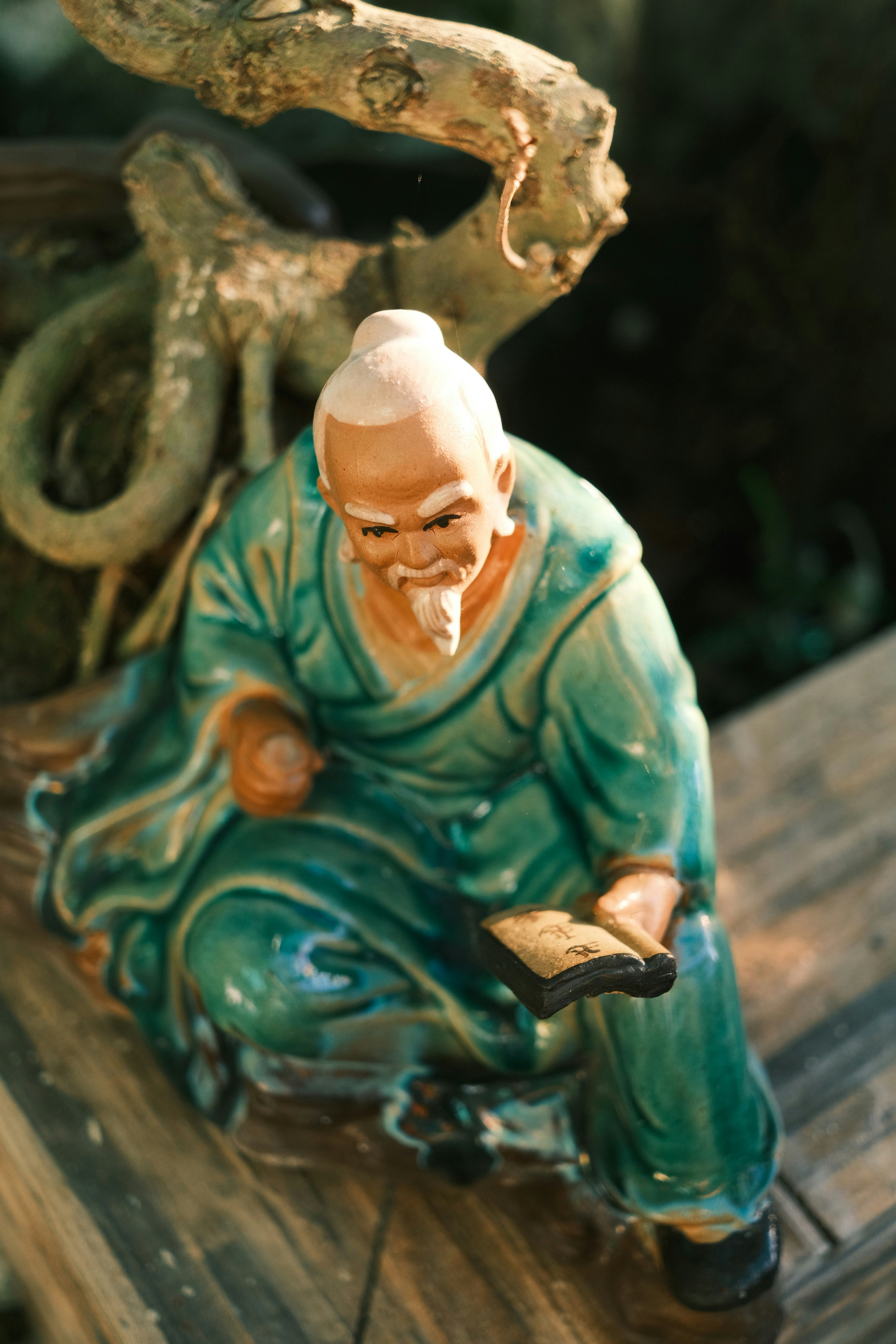 Ceramic figurine of an elderly scholar seated with a book, adorned in a flowing blue robe, surrounded by nature. The intricate details highlight the artistry of traditional craftsmanship.