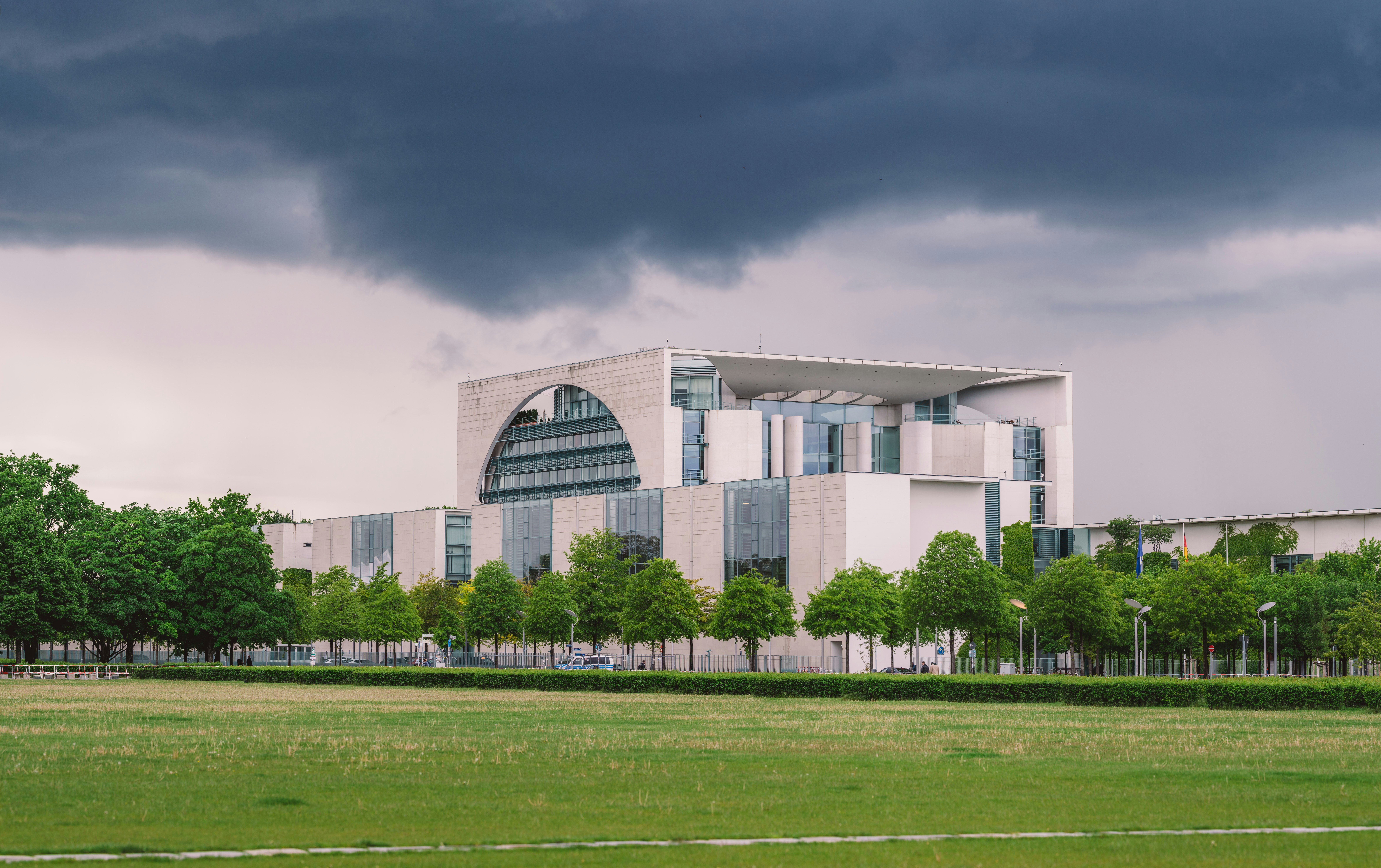 Modern government building with dramatic storm clouds overhead and lush greenery in the foreground.
