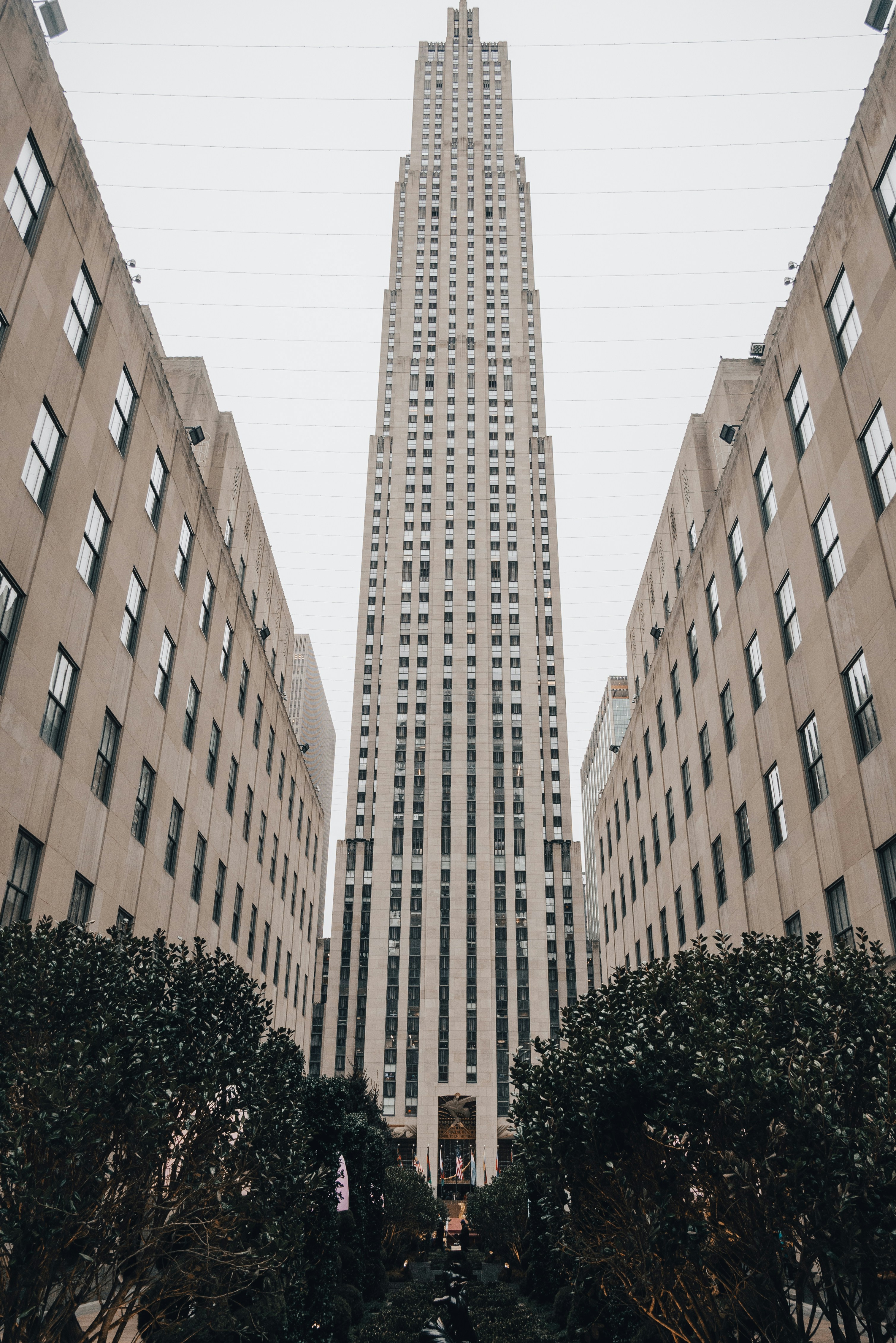 Art Deco skyscraper rises dramatically between symmetrical buildings, framed by lush greenery and a cloudy sky.