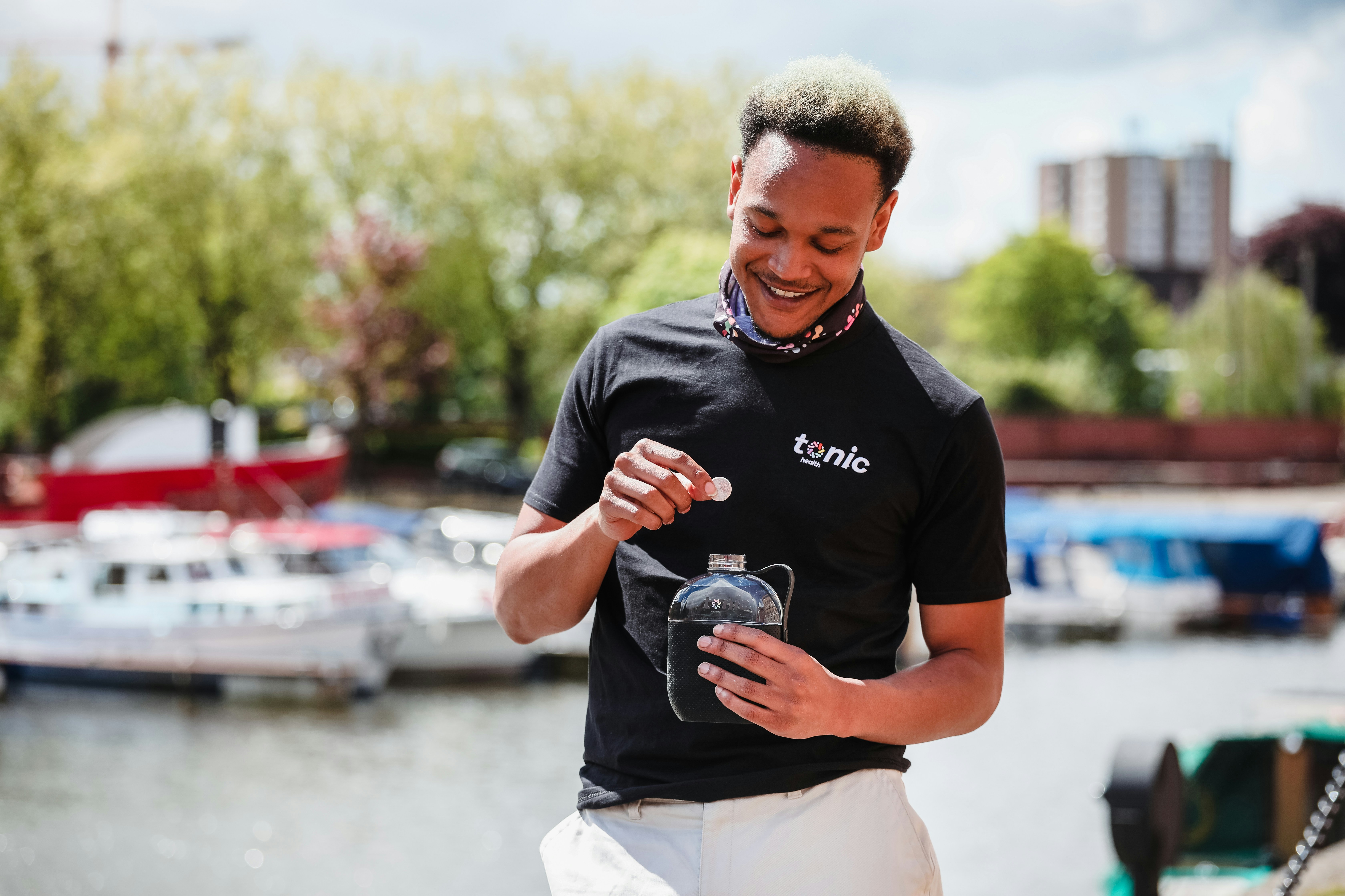 A young man in a black t-shirt smiles while holding a drink near a marina, surrounded by boats and greenery.