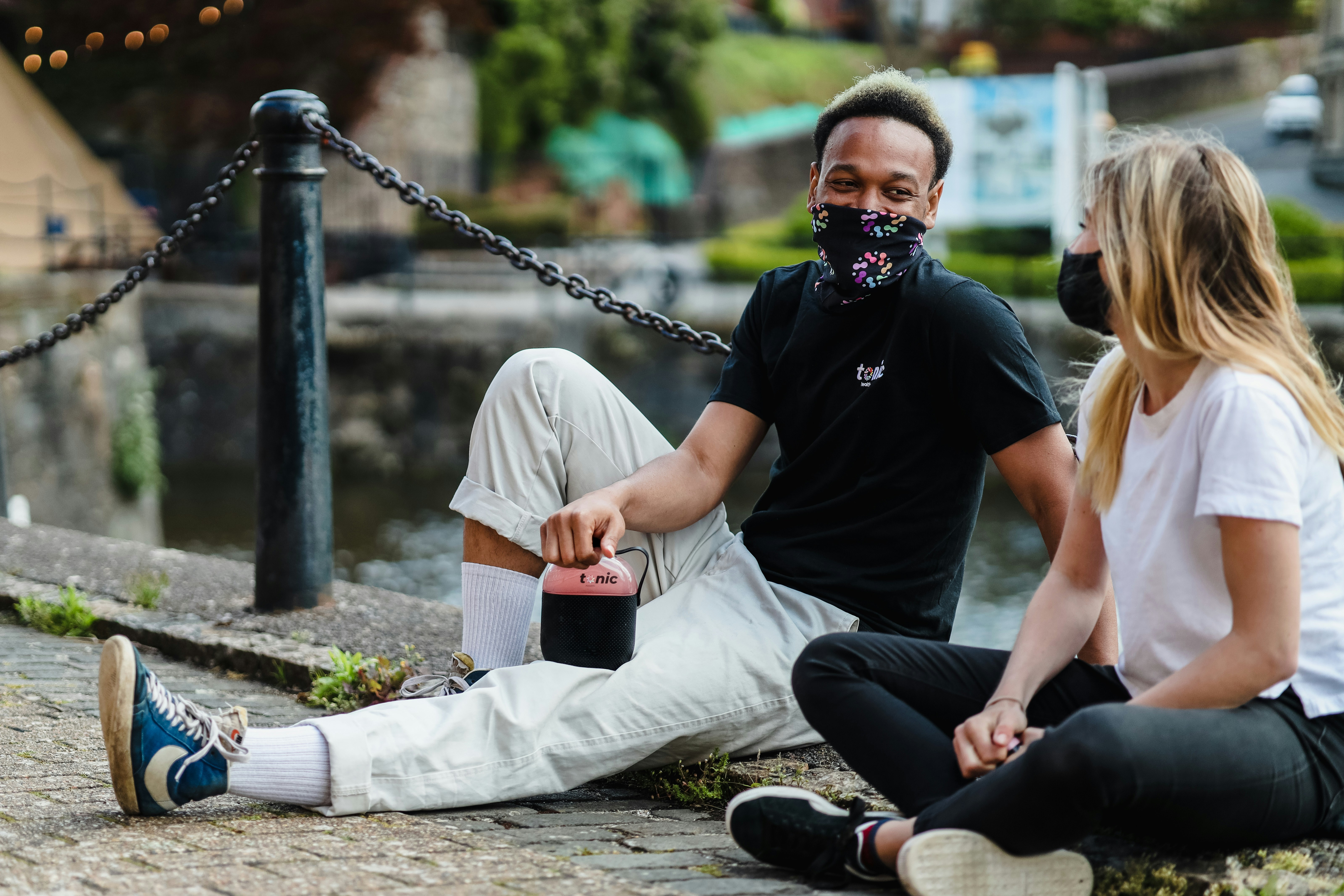 Two friends enjoying a relaxed moment by the water, engaged in conversation while wearing face masks. The setting features a scenic riverside backdrop.
