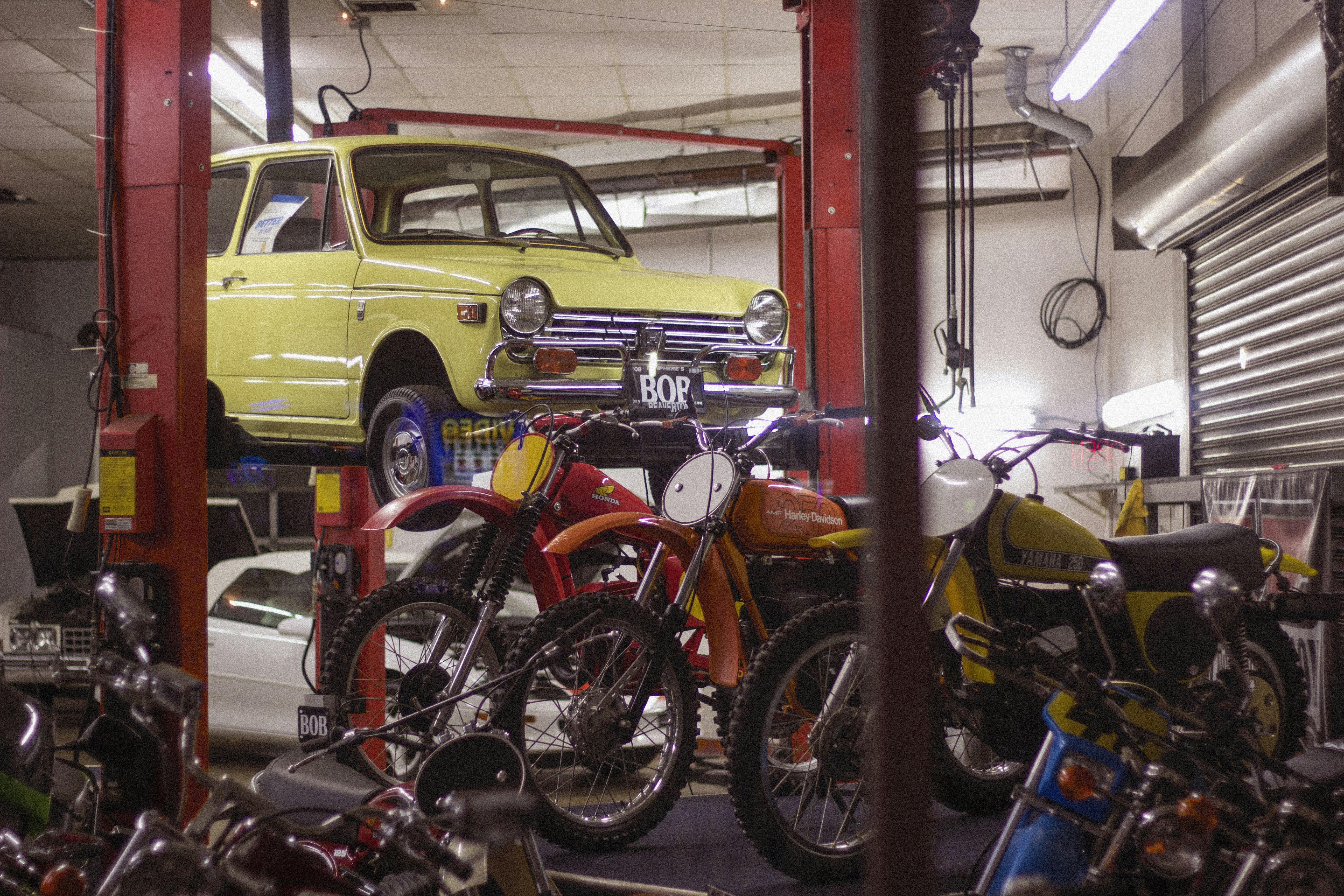 Yellow classic car perched on a lift above a clutter of vintage bikes in a busy workshop