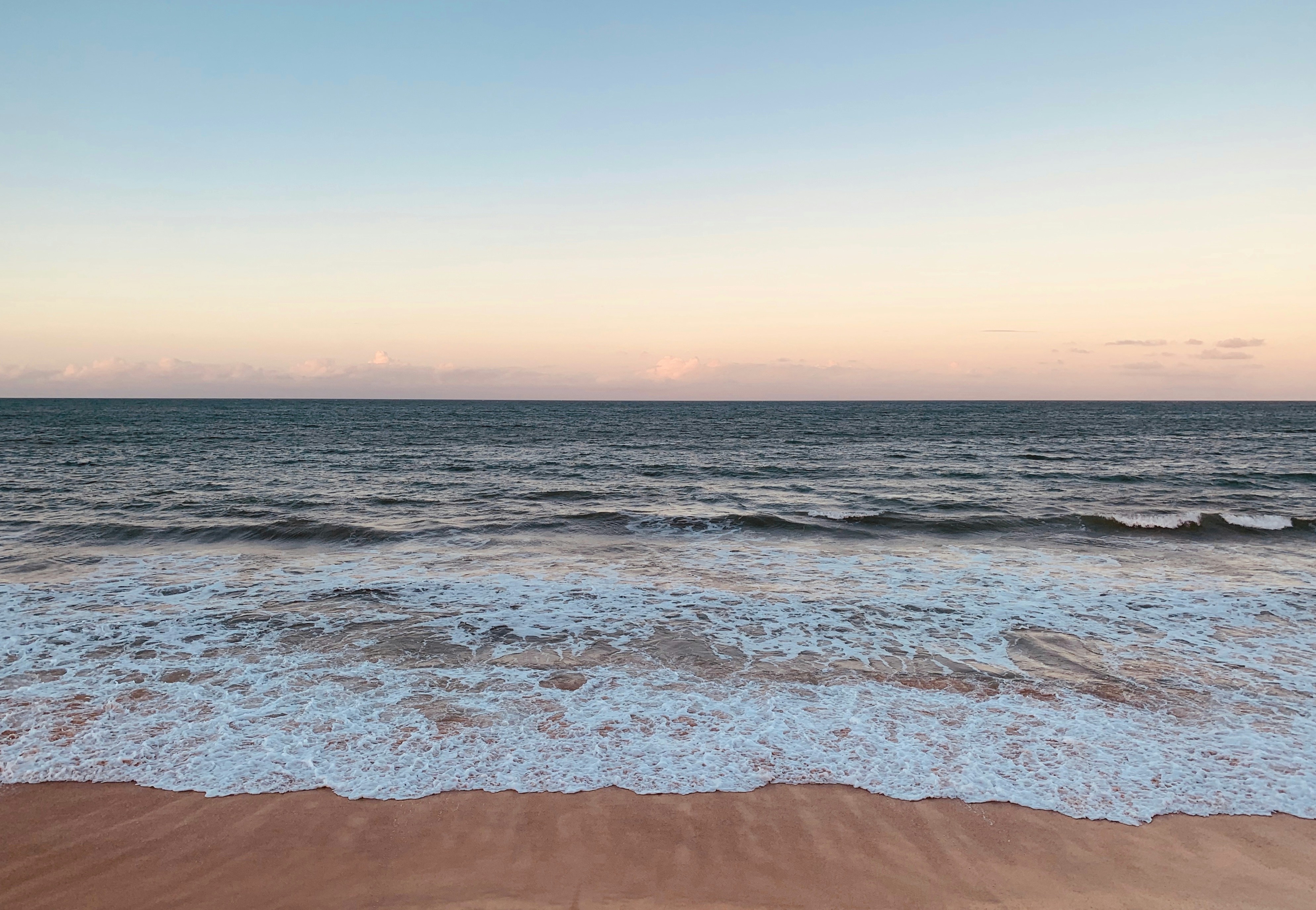 Gentle ocean waves roll onto a sandy beach under a pastel-hued sunset sky.