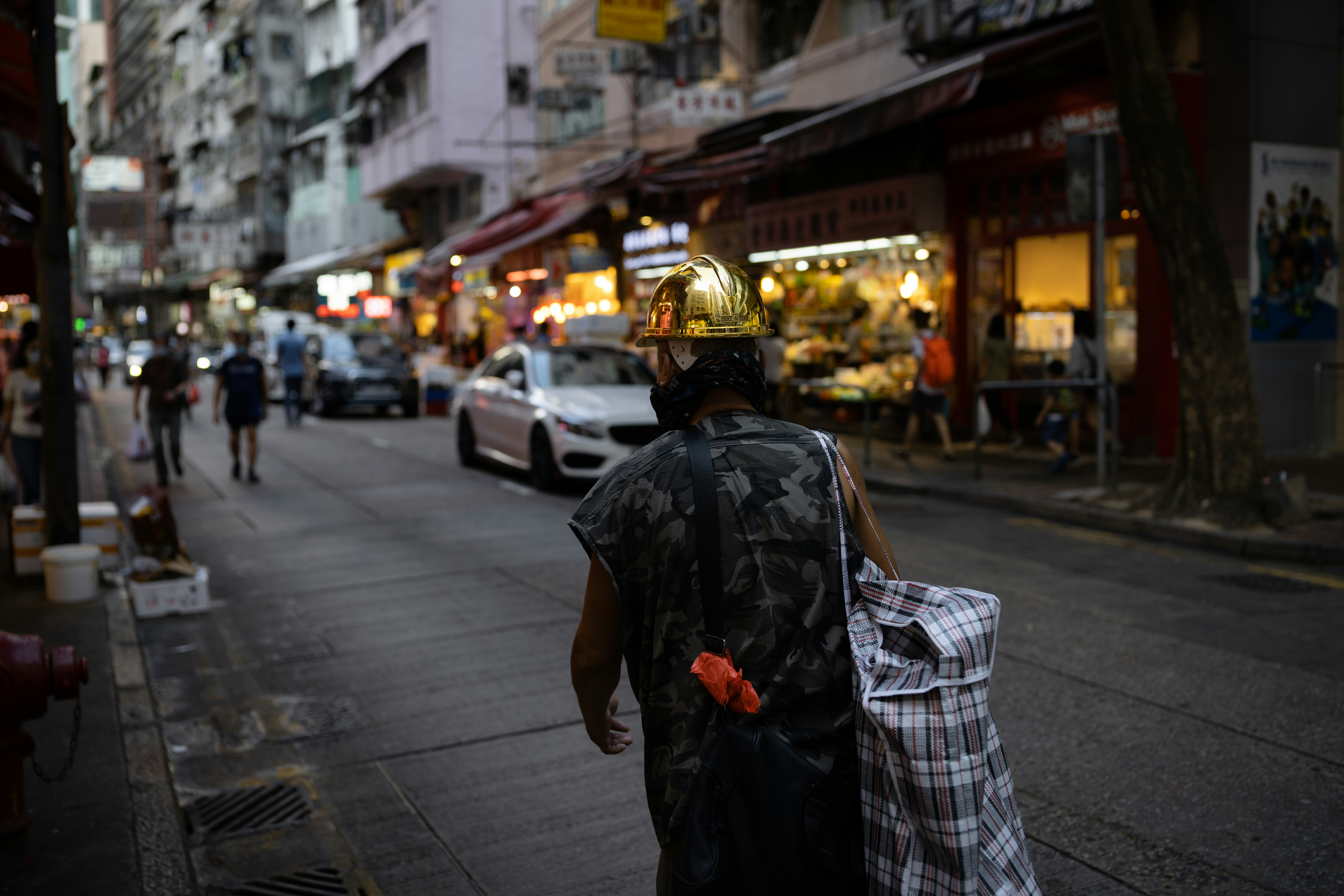 Person carrying a large checkered bag walks along a bustling urban street lined with glowing shopfronts at dusk.