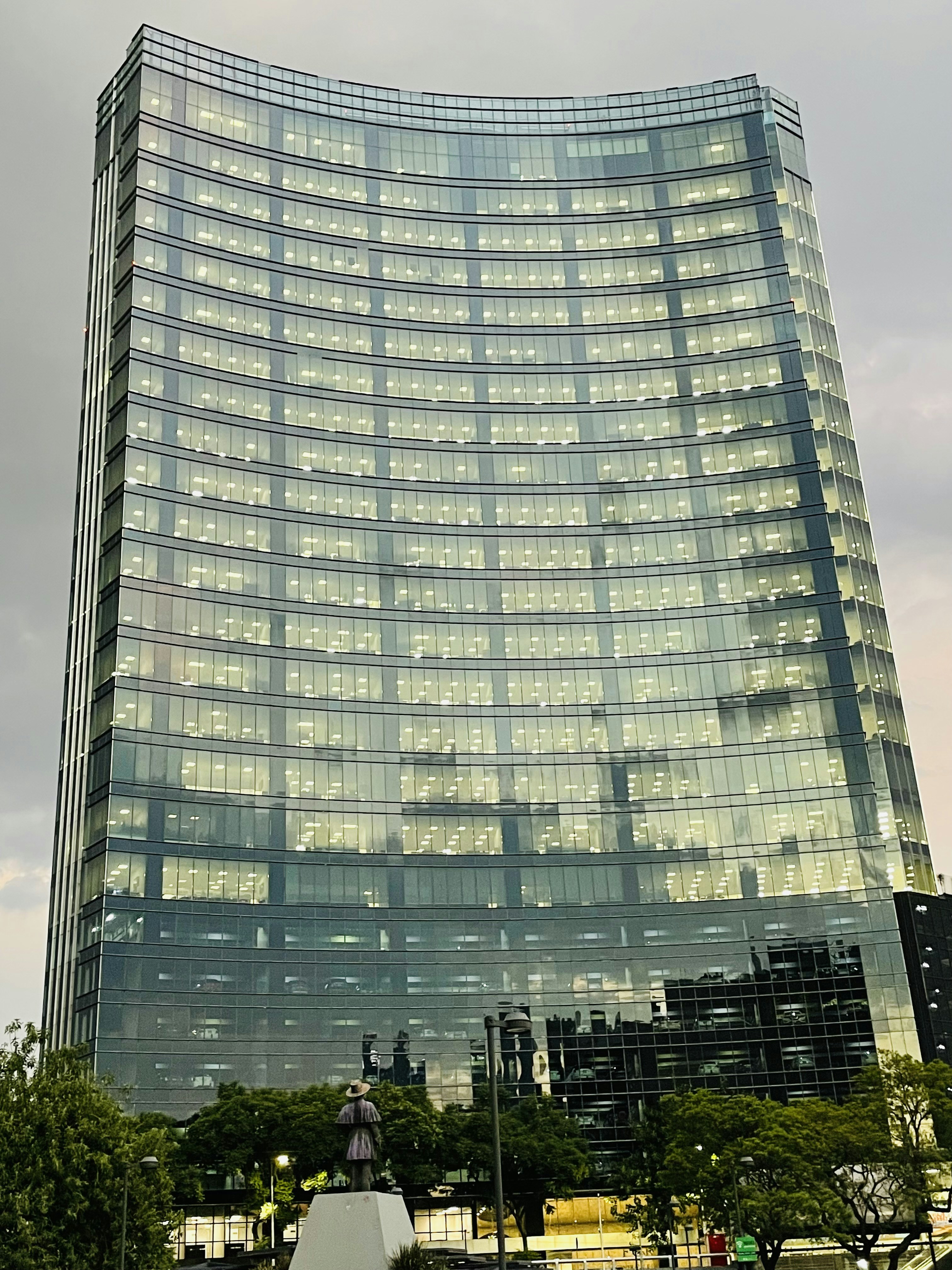 A sleek, curved glass building reflecting the surrounding urban landscape, with illuminated office spaces visible through the transparent exterior.