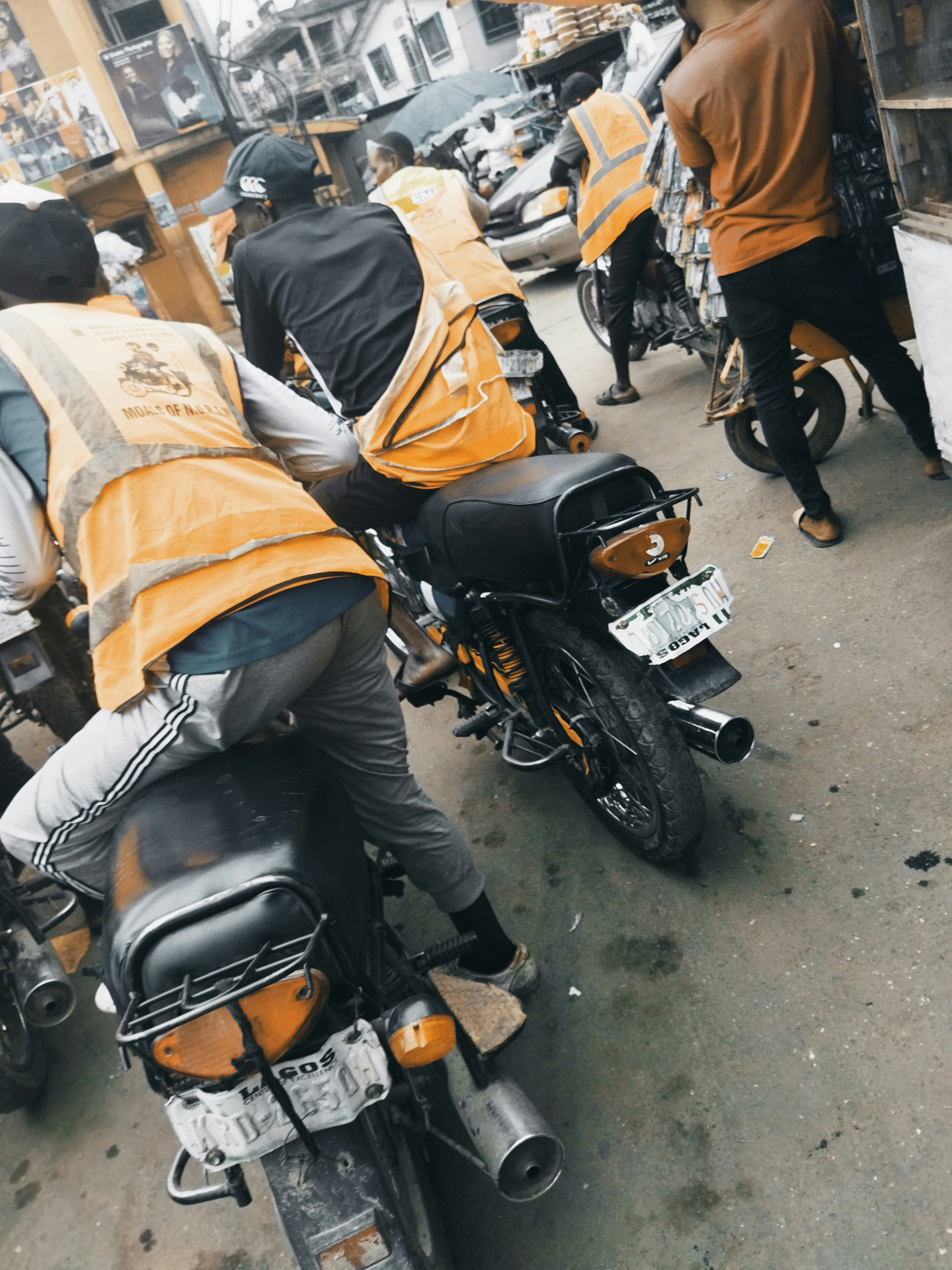 Crowded street scene with multiple motorbikes and riders in orange safety vests navigating a narrow urban street.