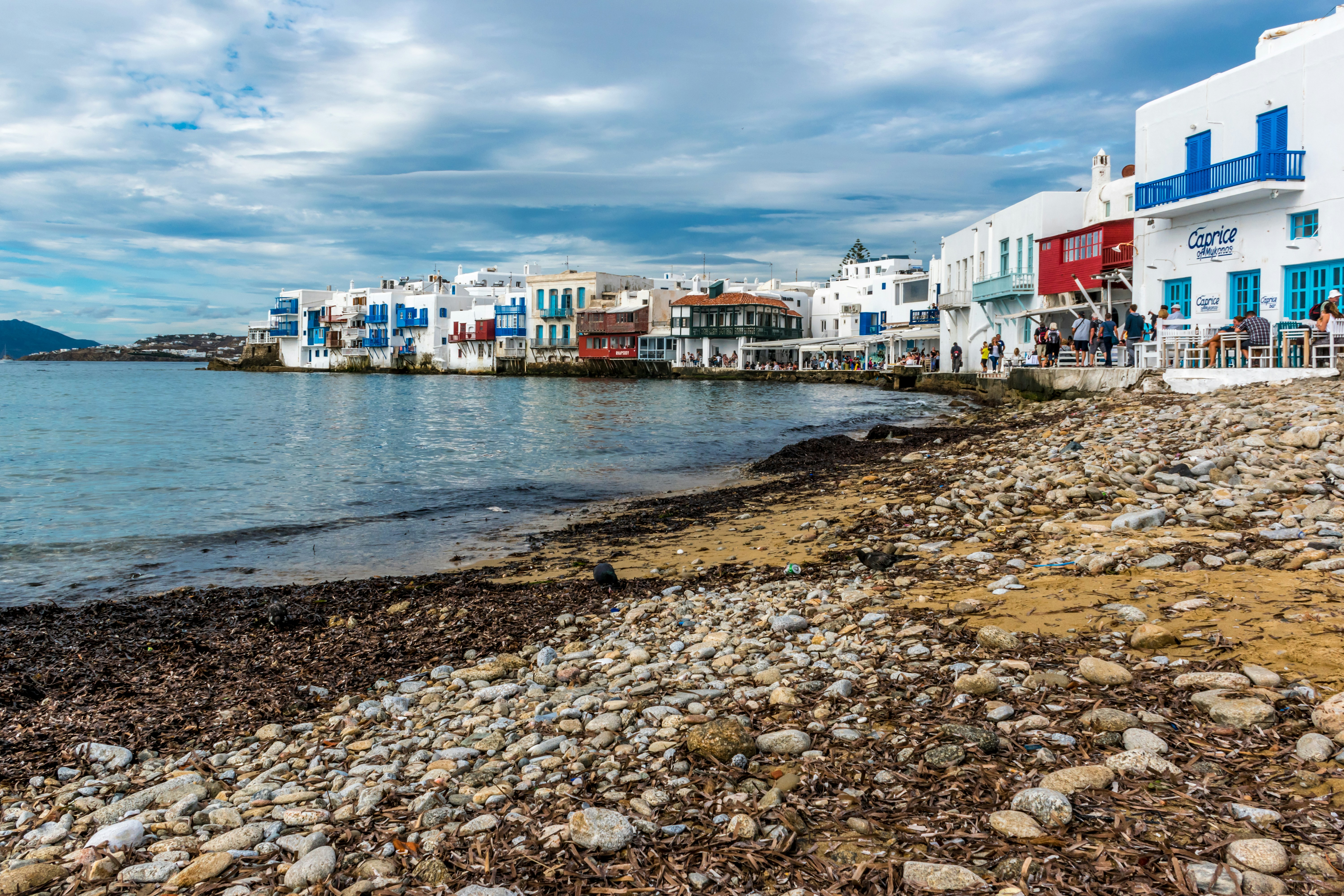 Pebbly shoreline leading to colorful buildings along the Mykonos waterfront under a dynamic sky.