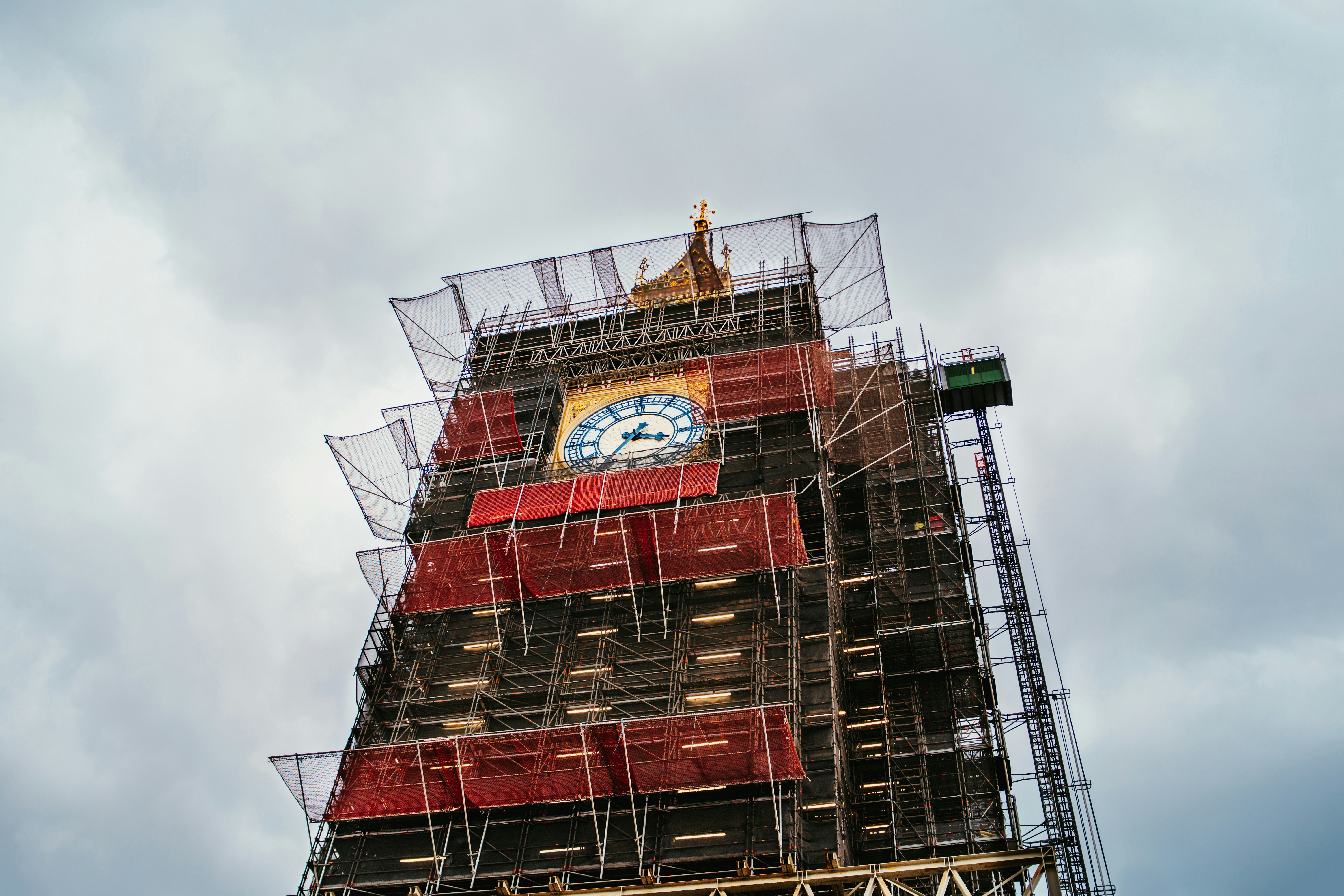 This striking image captures the famous clock tower enveloped in scaffolding, set against a backdrop of moody, overcast skies. The composition highlights the intricate network of red and silver scaffolding that contrasts with the face of the clock and its golden accents. The soft, diffused lighting enhances the somber atmosphere, while the muted color palette brings attention to the ongoing restoration work, making it a visually captivating scene.