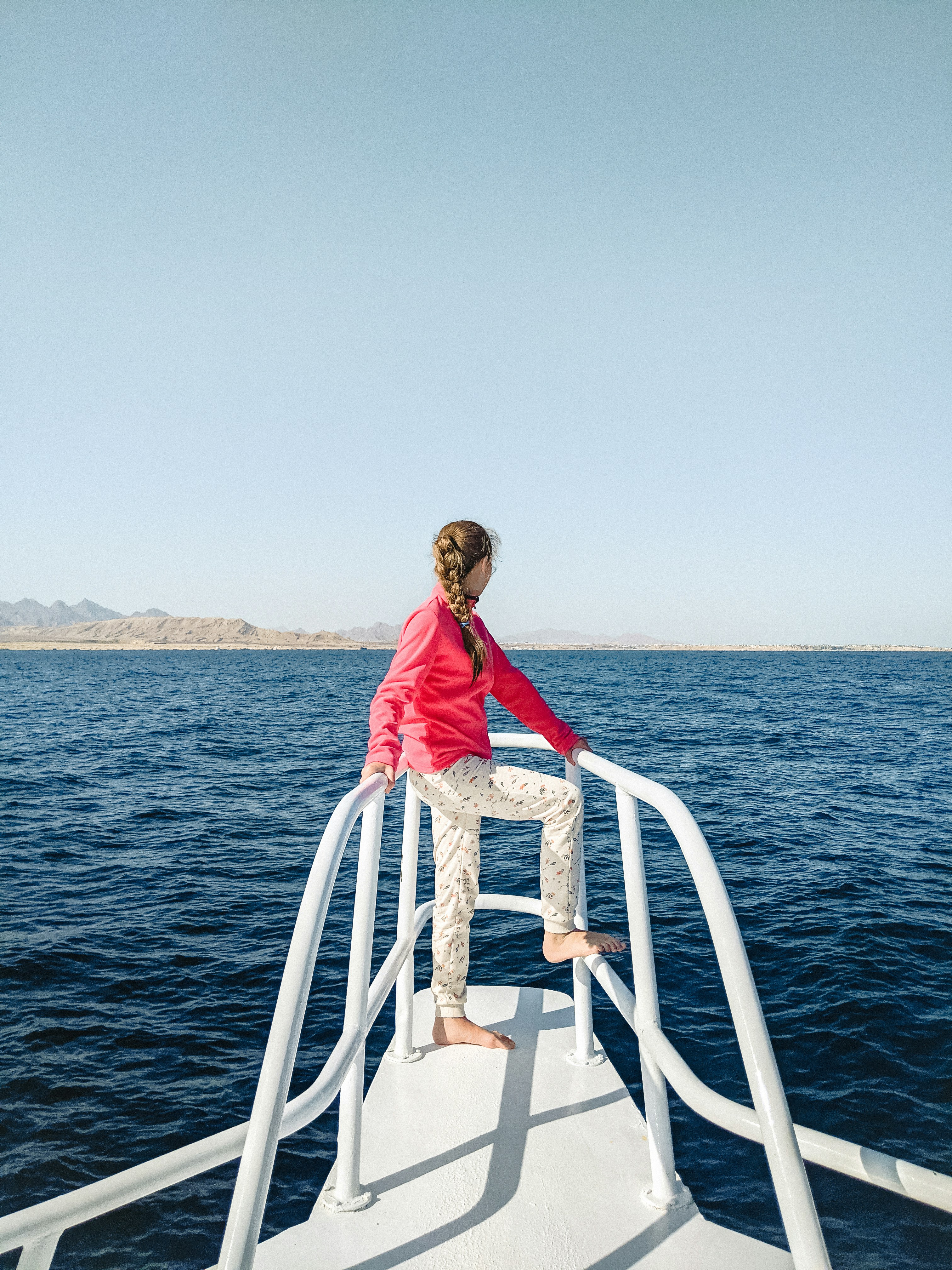 A girl with braided hair in a red jacket stands barefoot on the bow of a boat, facing the open ocean. The scene highlights vast blue water, distant land on the horizon, and a calm, sunlit sky.
