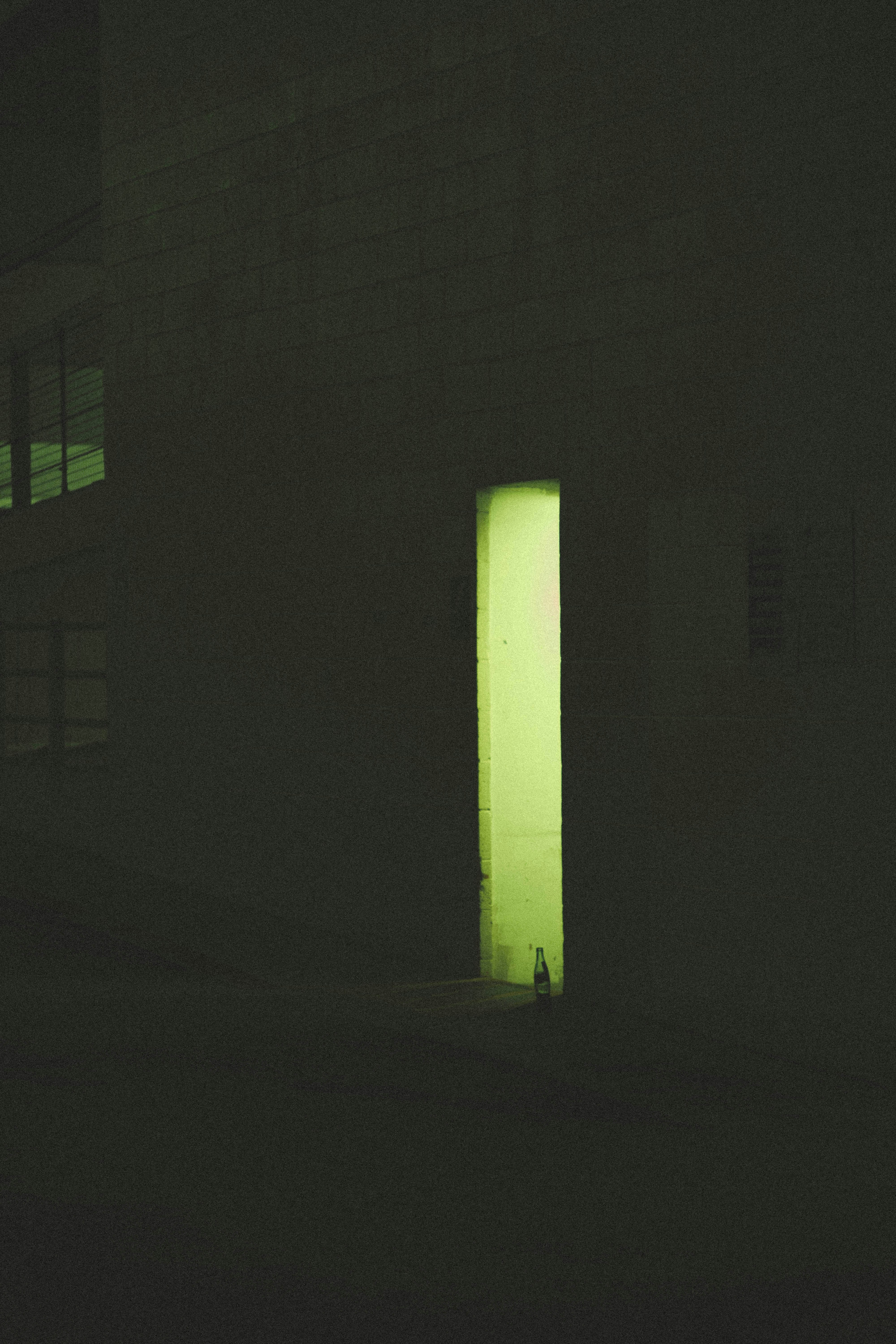 A faintly illuminated green door stands ajar against a dark, shadowy building, hinting at secrets within.