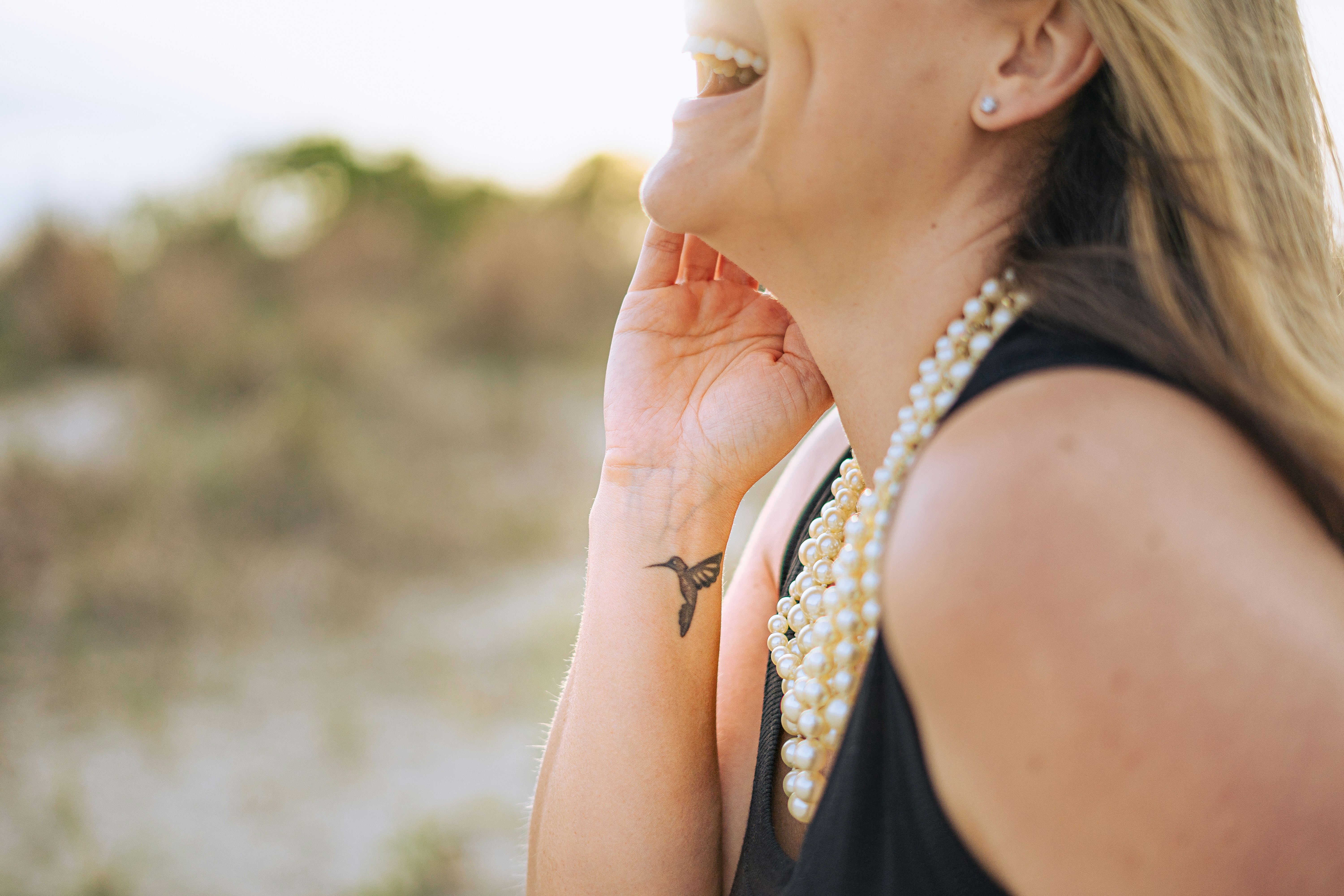 Woman laughing with a pearl necklace, showcasing a tattoo on her wrist, against a blurred natural background.