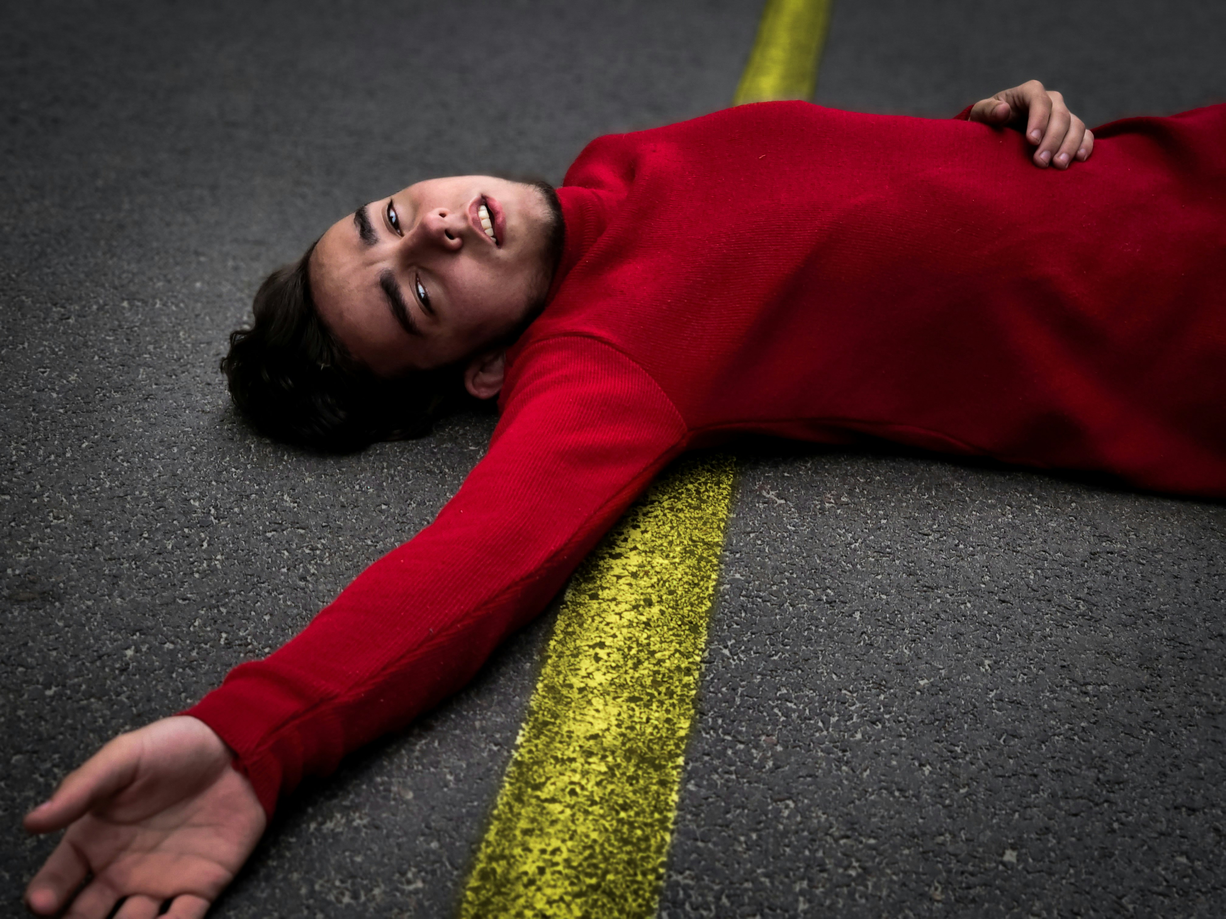A young man in a red sweater lies on the ground, arms outstretched beside a yellow line, evoking a sense of vulnerability and introspection.
